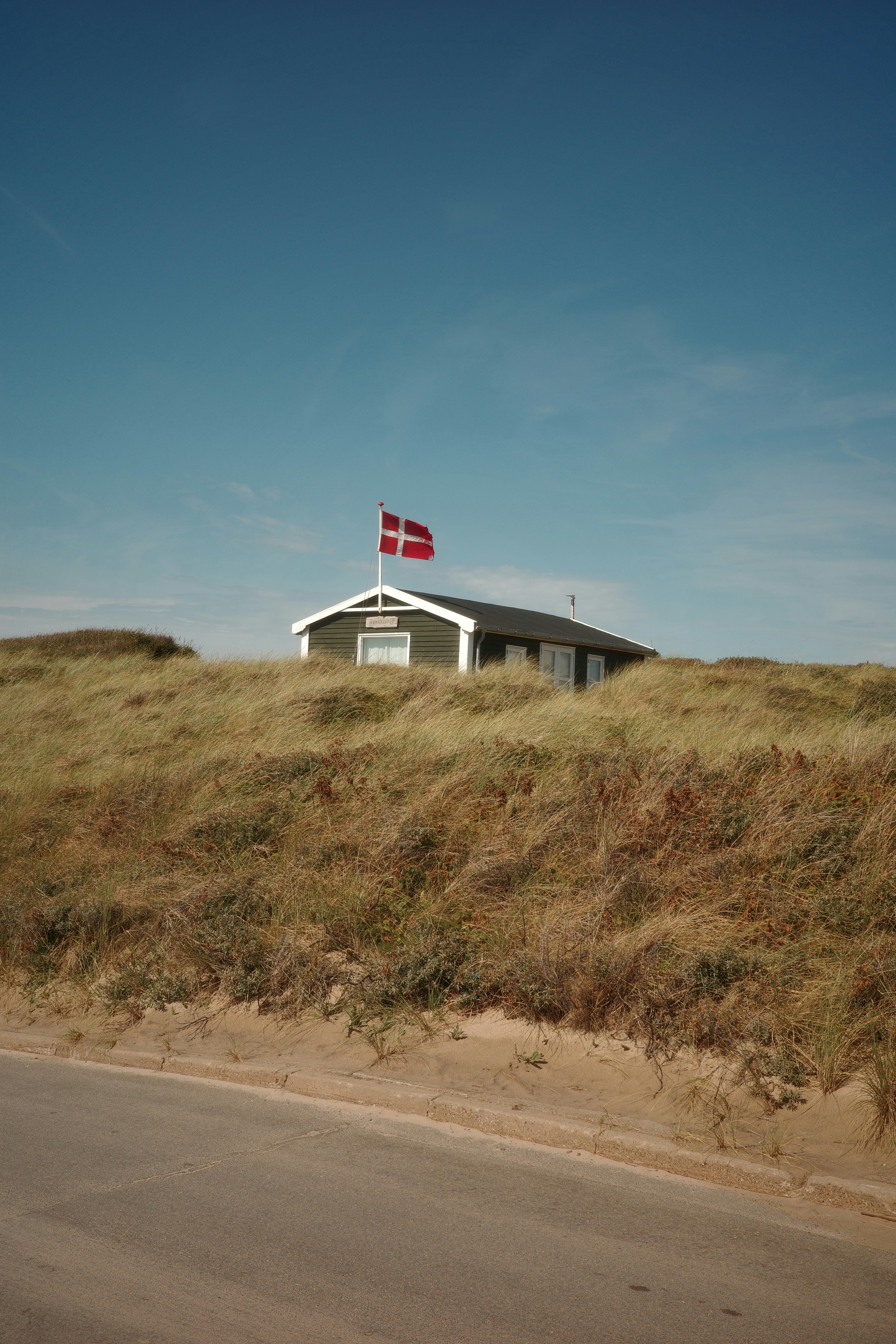 Small coastal house with a Danish flag sits atop grassy dunes under a clear blue sky.