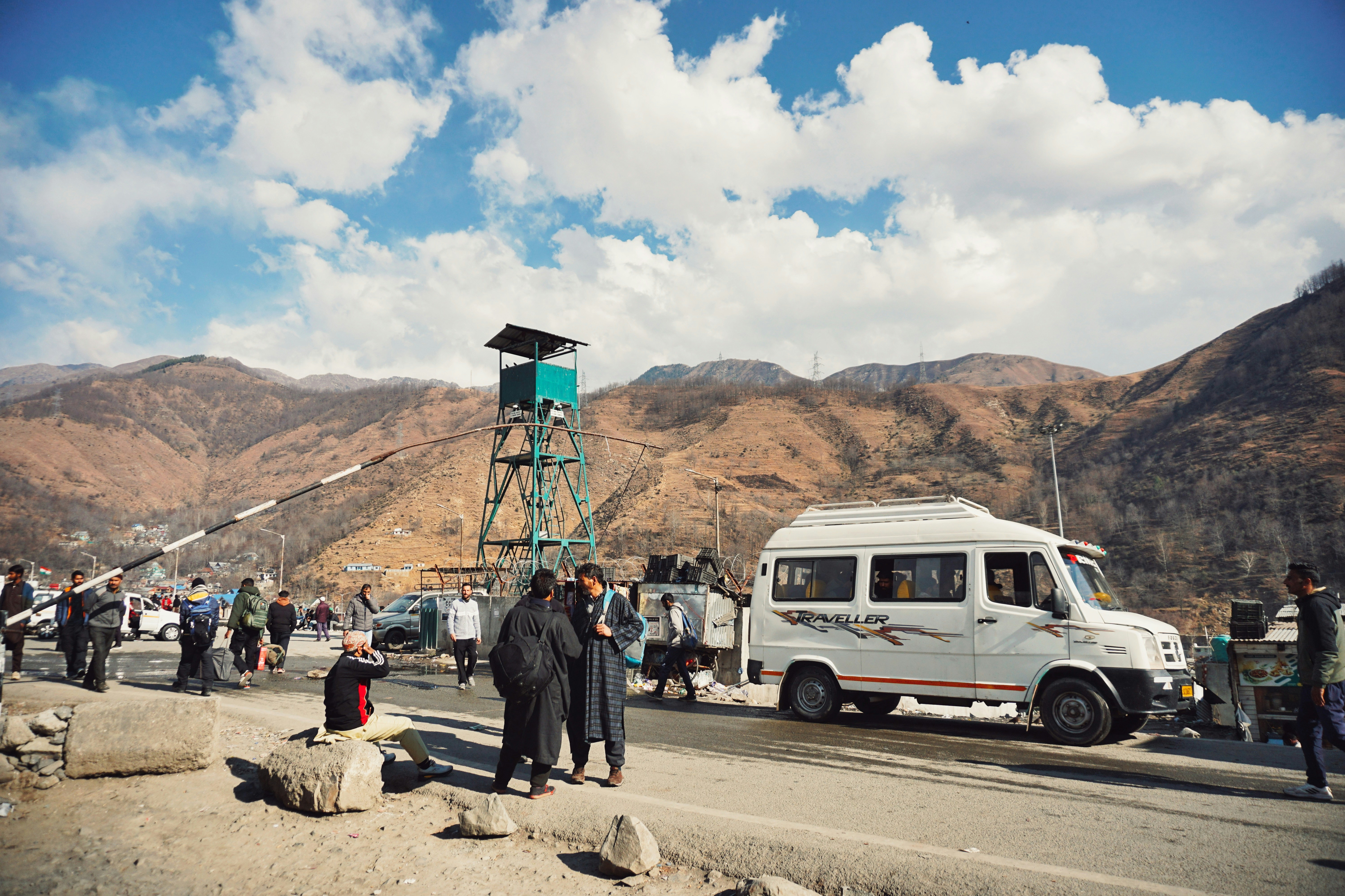 A border checkpoint scene with a tall watchtower, a white van, and travelers gathered near rocky terrain under a bright, partly cloudy sky.