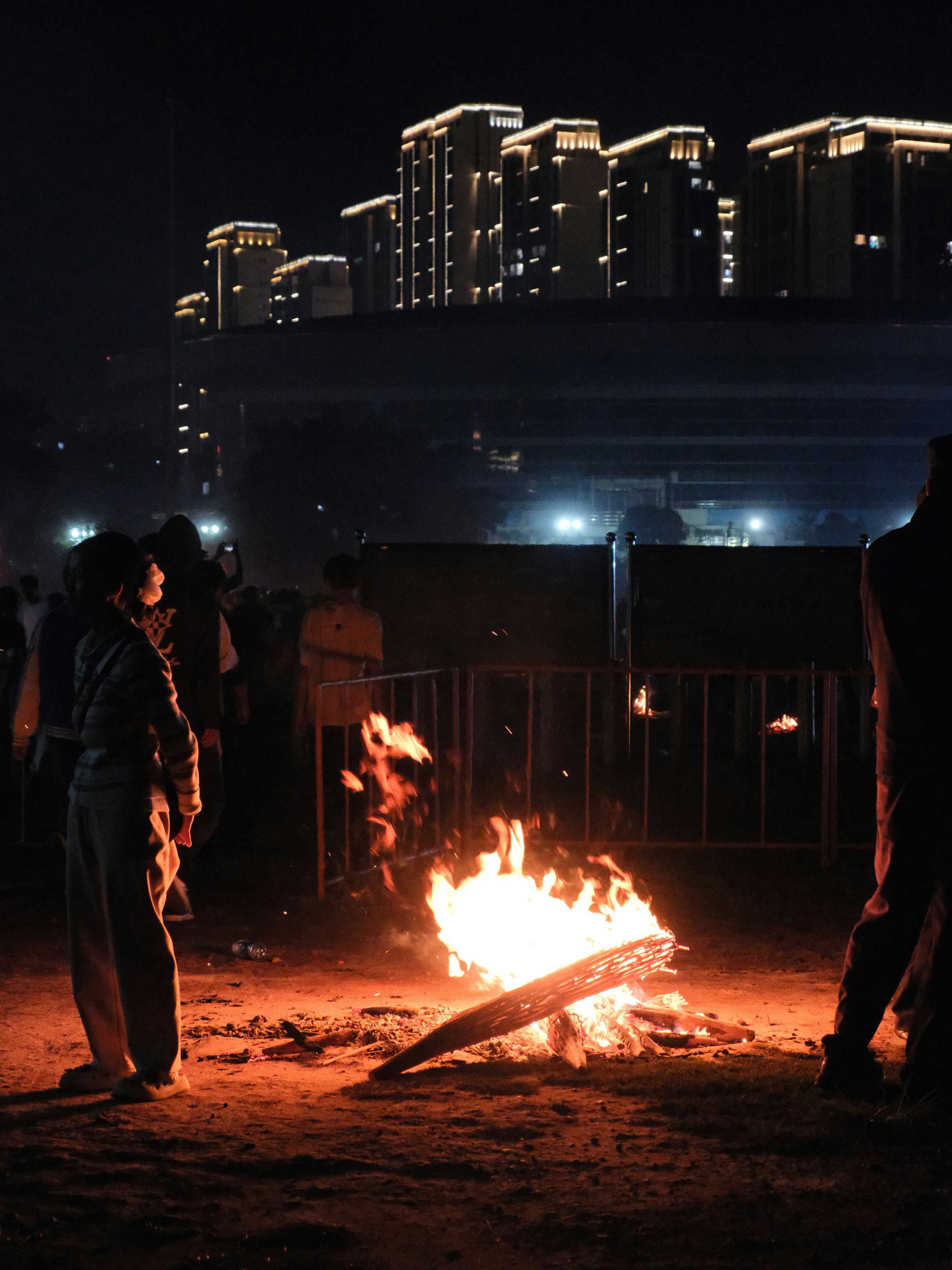 A nocturnal scene of a curling campfire in the foreground with silhouetted figures and a line of illuminated city towers in the background, set under a dark sky. It captures a communal moment against an urban skyline.