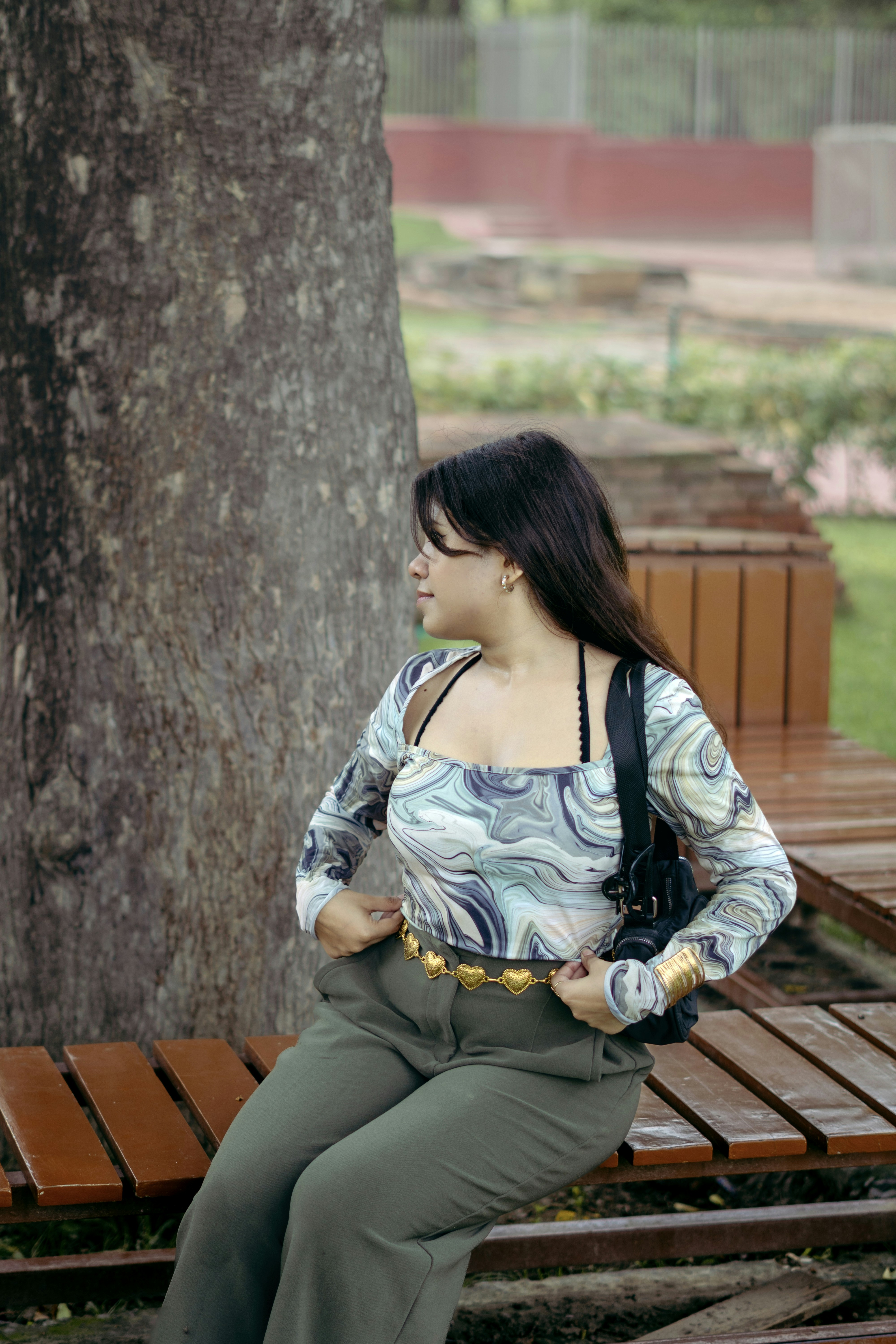A woman sits on a wooden bench beside a large tree, wearing a patterned top and olive pants with a gold belt, holding a camera bag in the park.