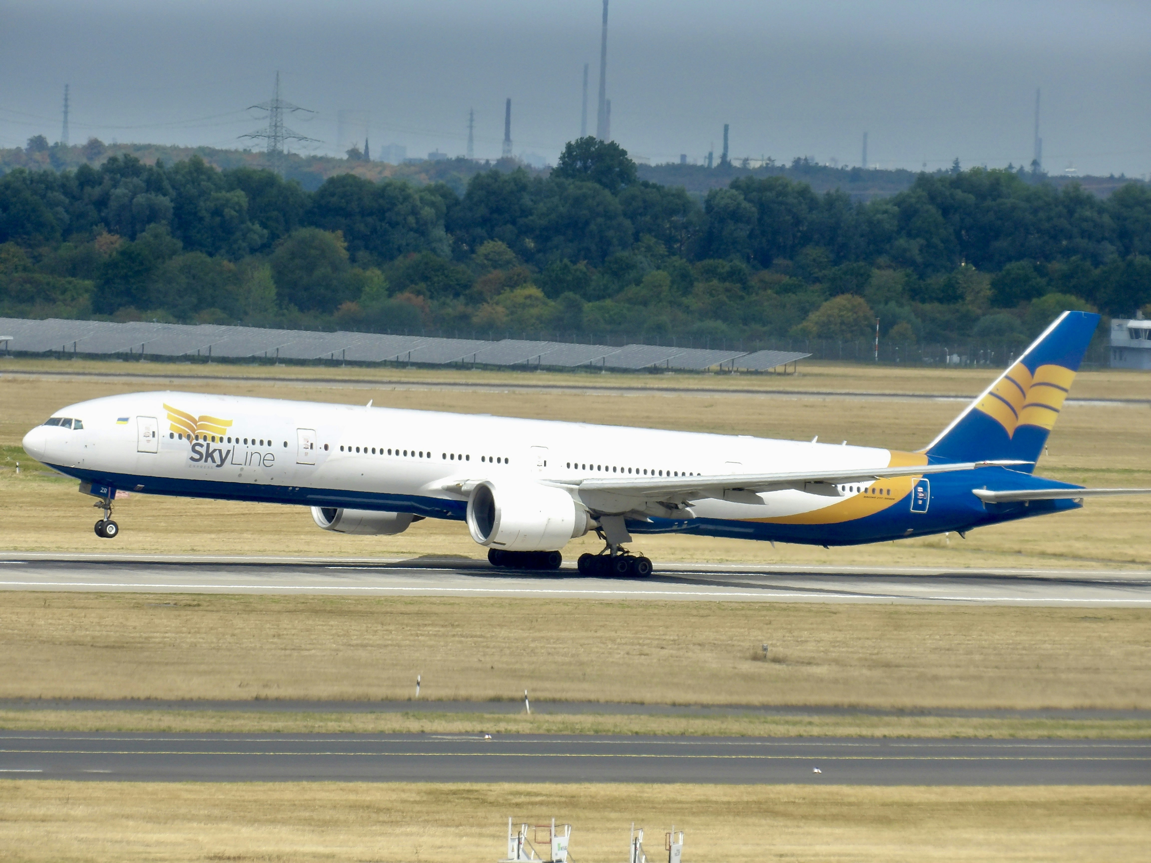 Airliner taxis along the runway with a distant tree line and industrial backdrop visible in the background. The scene captures the subject in motion, emphasizing the aircraft's form and livery.