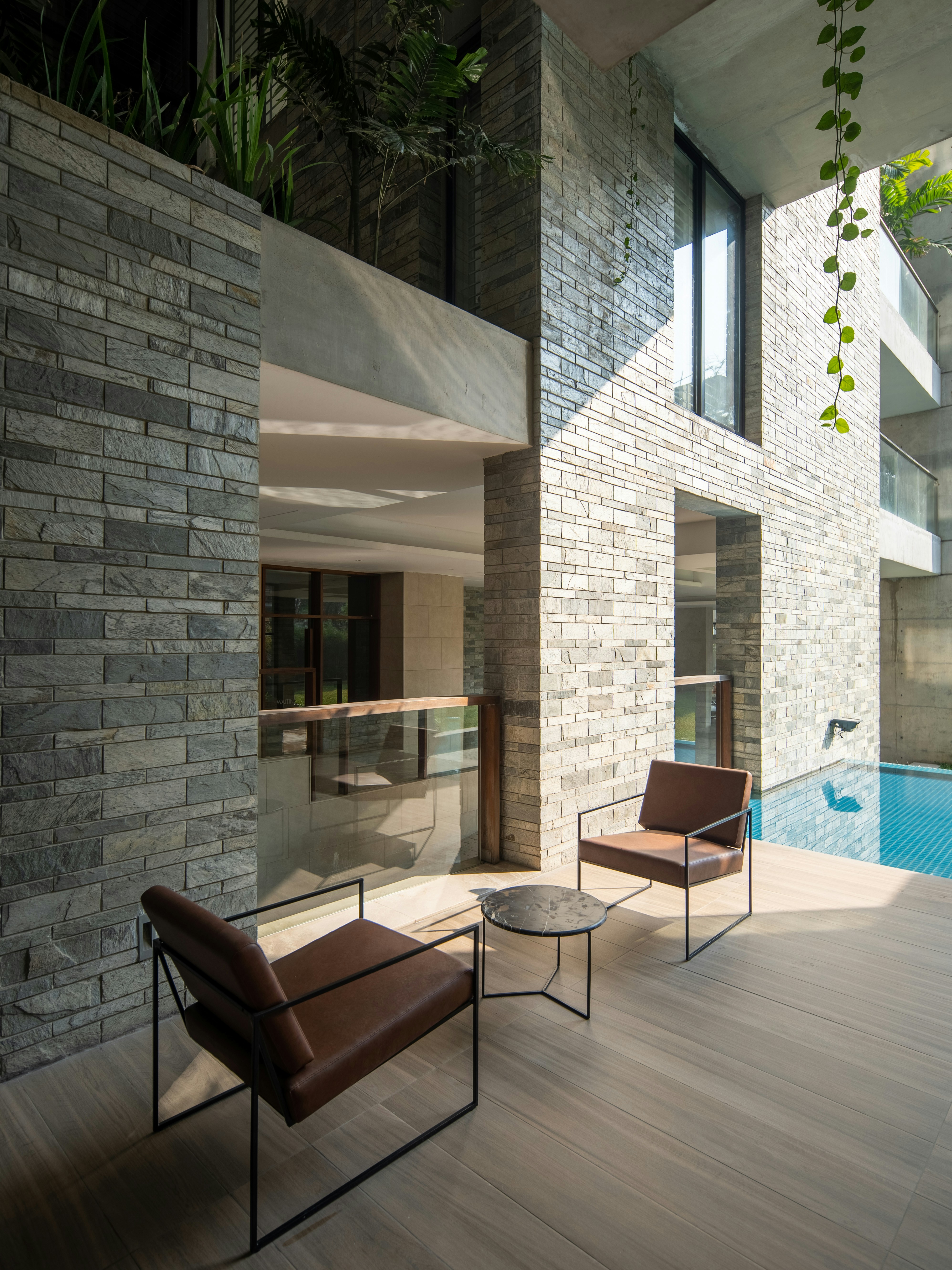 Contemporary outdoor lounge area with brown leather chairs and a small round table along a stone-clad wall beside a pool.