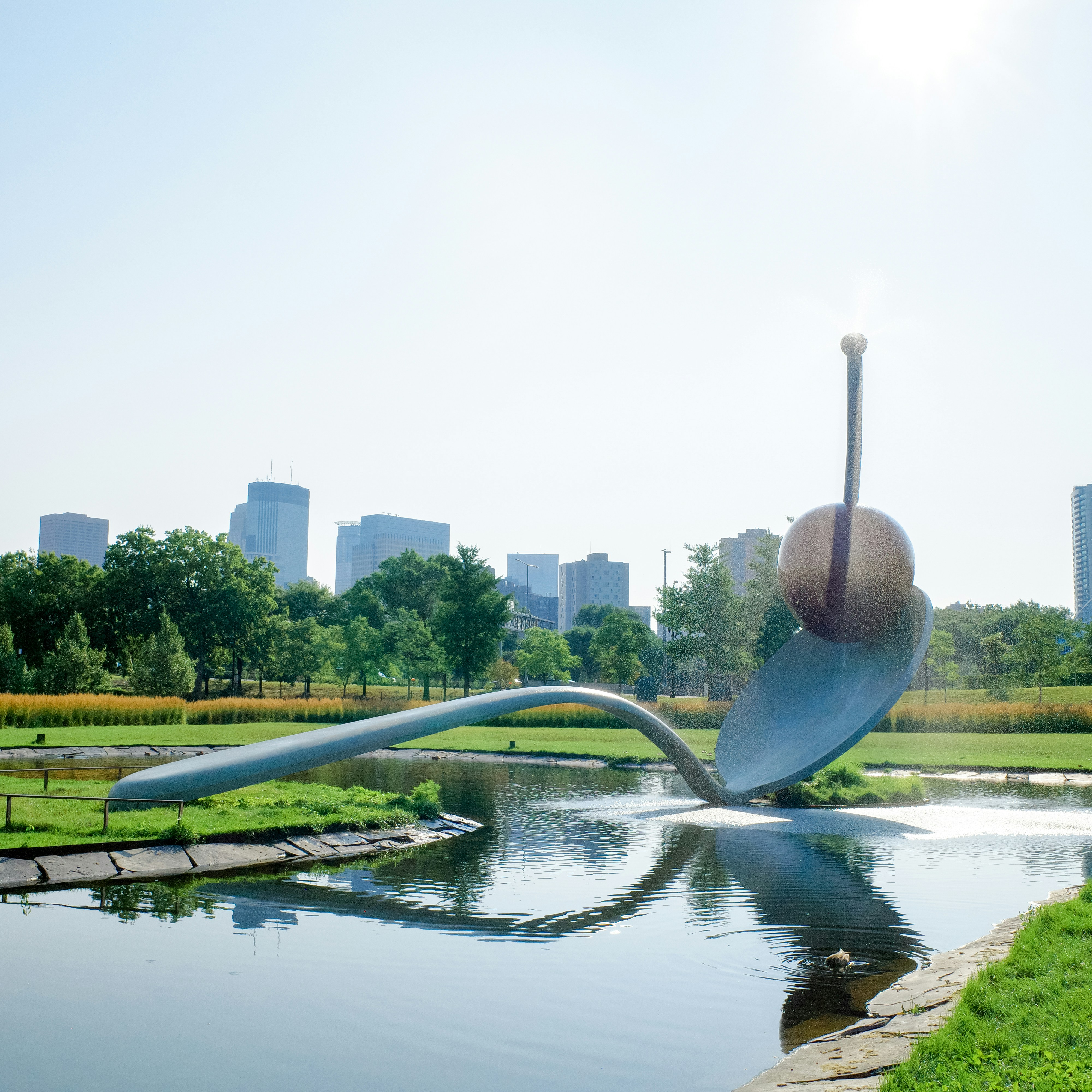 Sculptural metal arc rises from a tranquil reflecting pool with a spherical top, set against a green park and distant skyline.