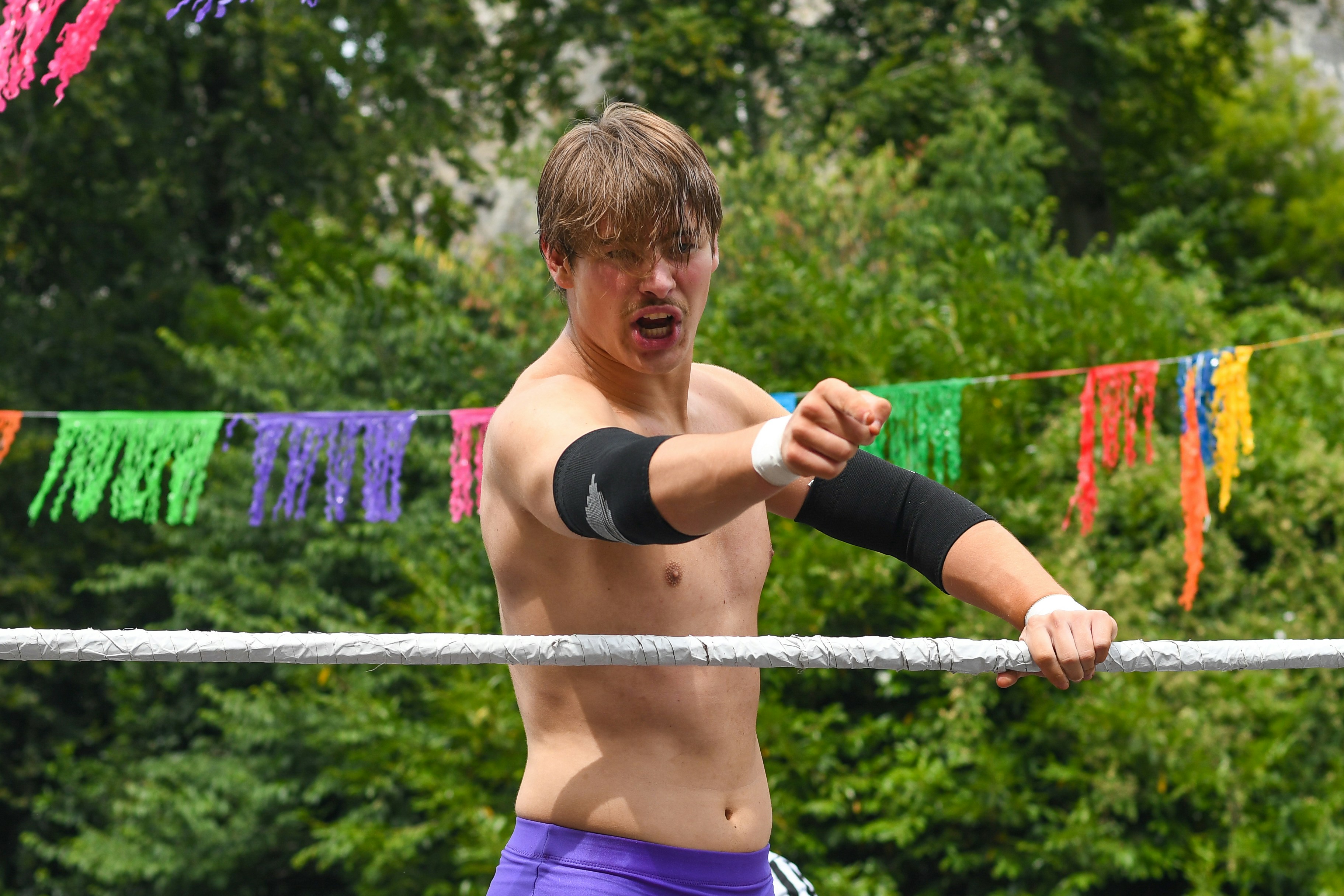 Energetic wrestler mid-action with right fist forward in an outdoor ring, colorful pennants and green backdrop providing contrast.