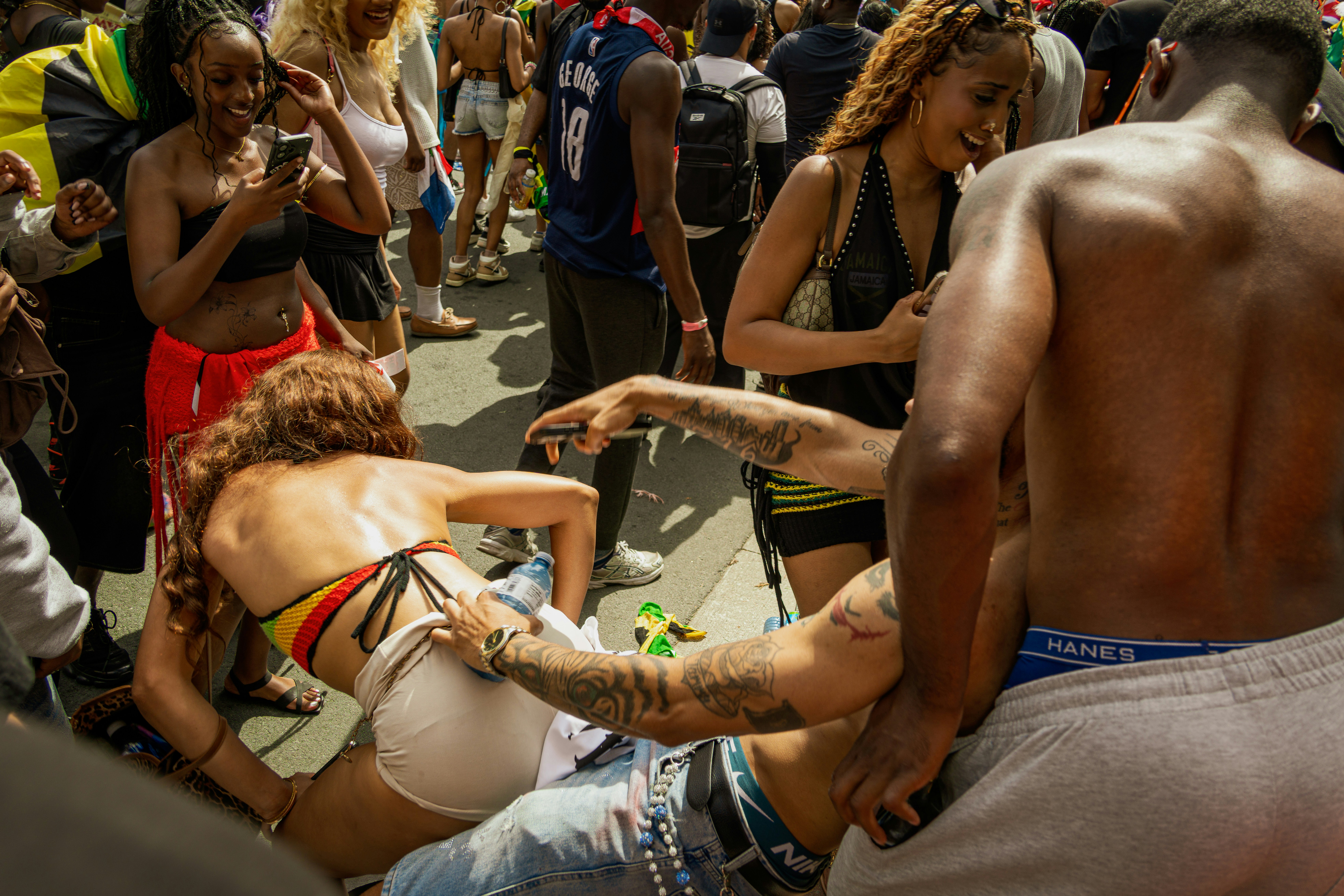 Energetic crowd gathering around a dancer in swimwear, with a tattooed arm reaching toward a performer in the foreground at a sunny outdoor party.