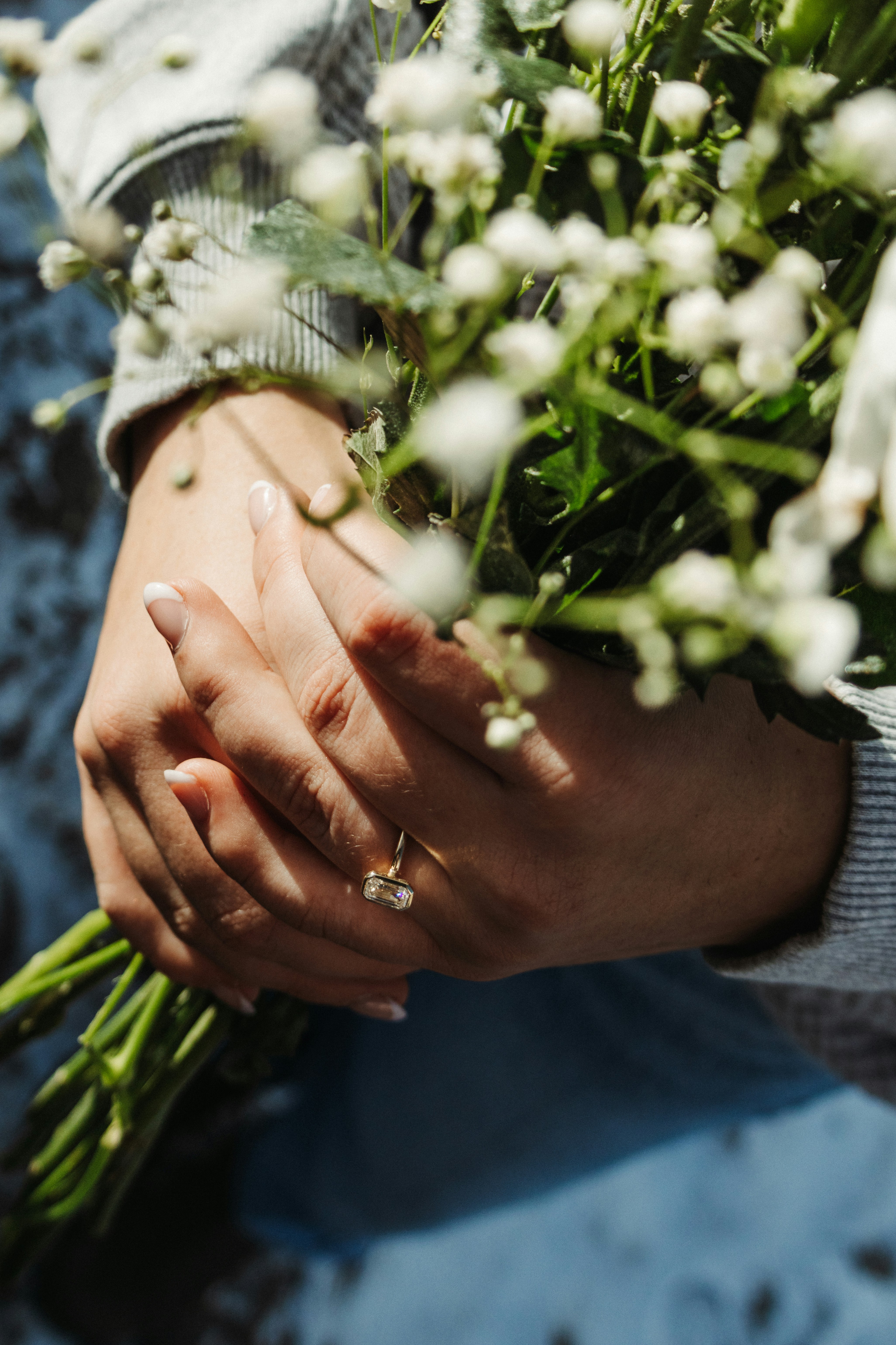 A close-up of intertwined hands holding a bouquet of white blossoms, emphasizing a ring and soft skin tones; a candid, intimate moment with a hint of greenery and casual clothing.