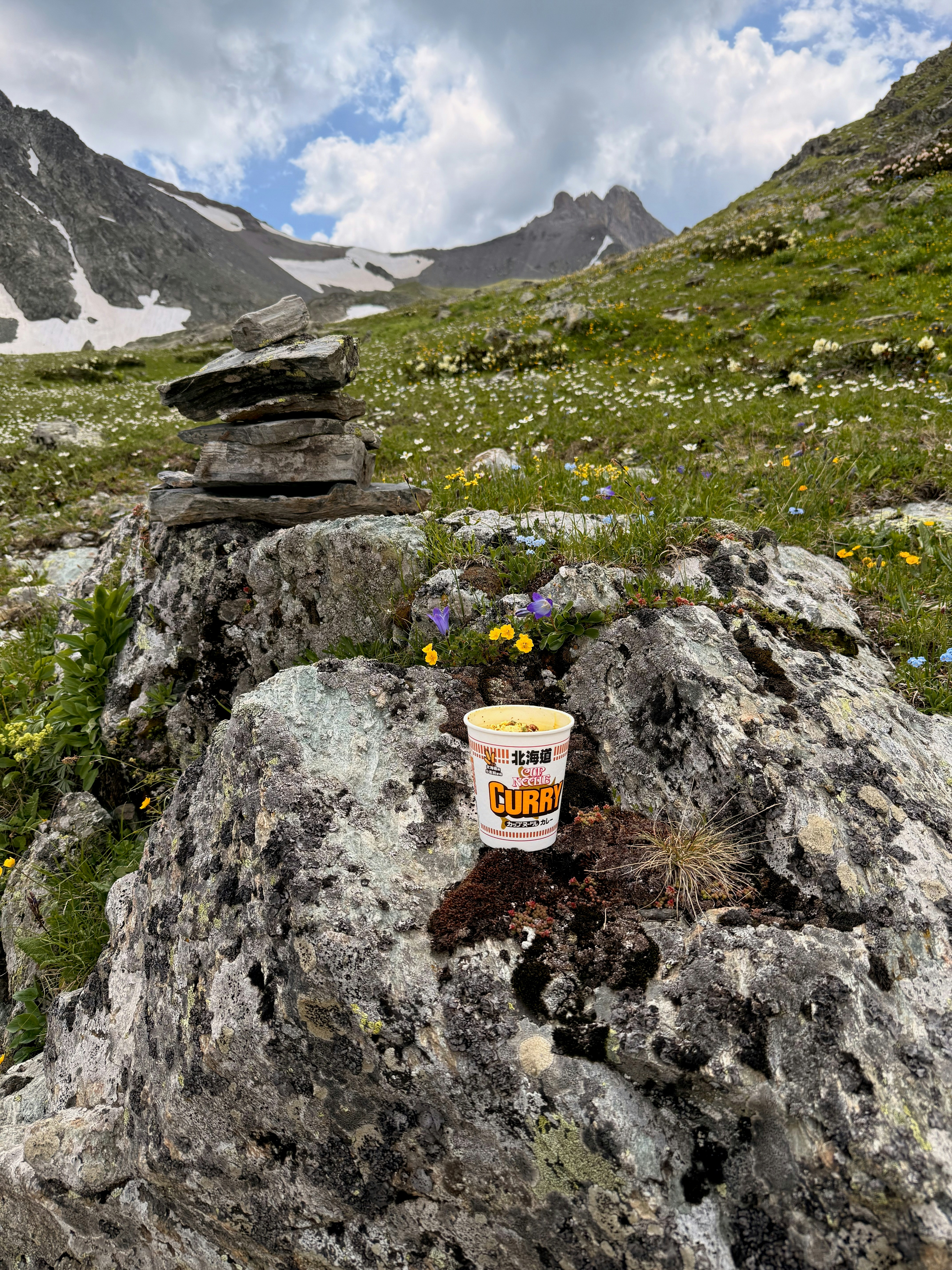 A cup of instant noodles on a mossy rock beside a stone cairn in mountains