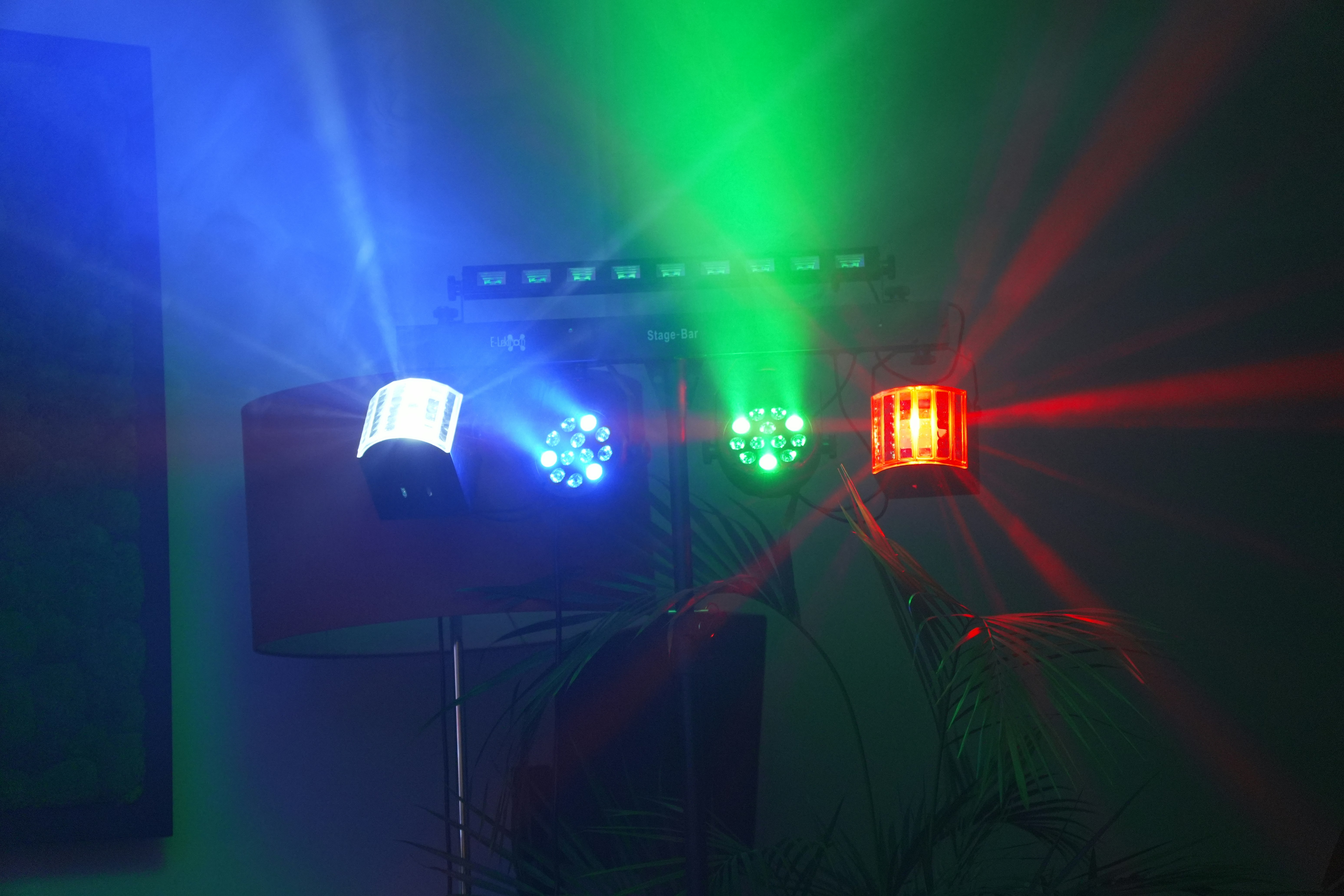 Multicolored LED stage lights emit blue, green, and red beams in a dim setting, with a metal stand and foliage hinting at an indoor studio or performance space.
