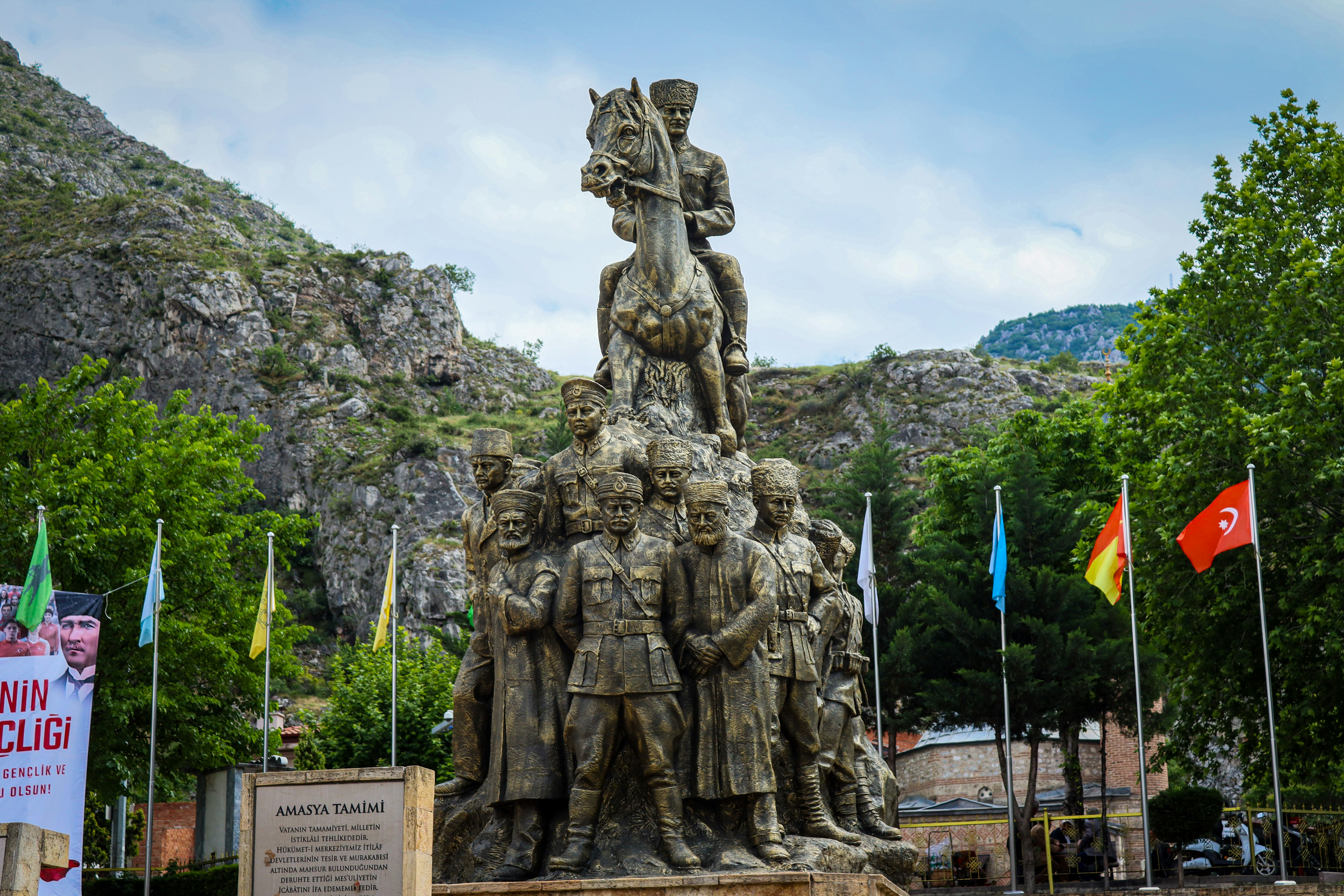 Monumental statue depicting clustered soldiers and a horse-mounted figure, flanked by colorful flags in a public square.