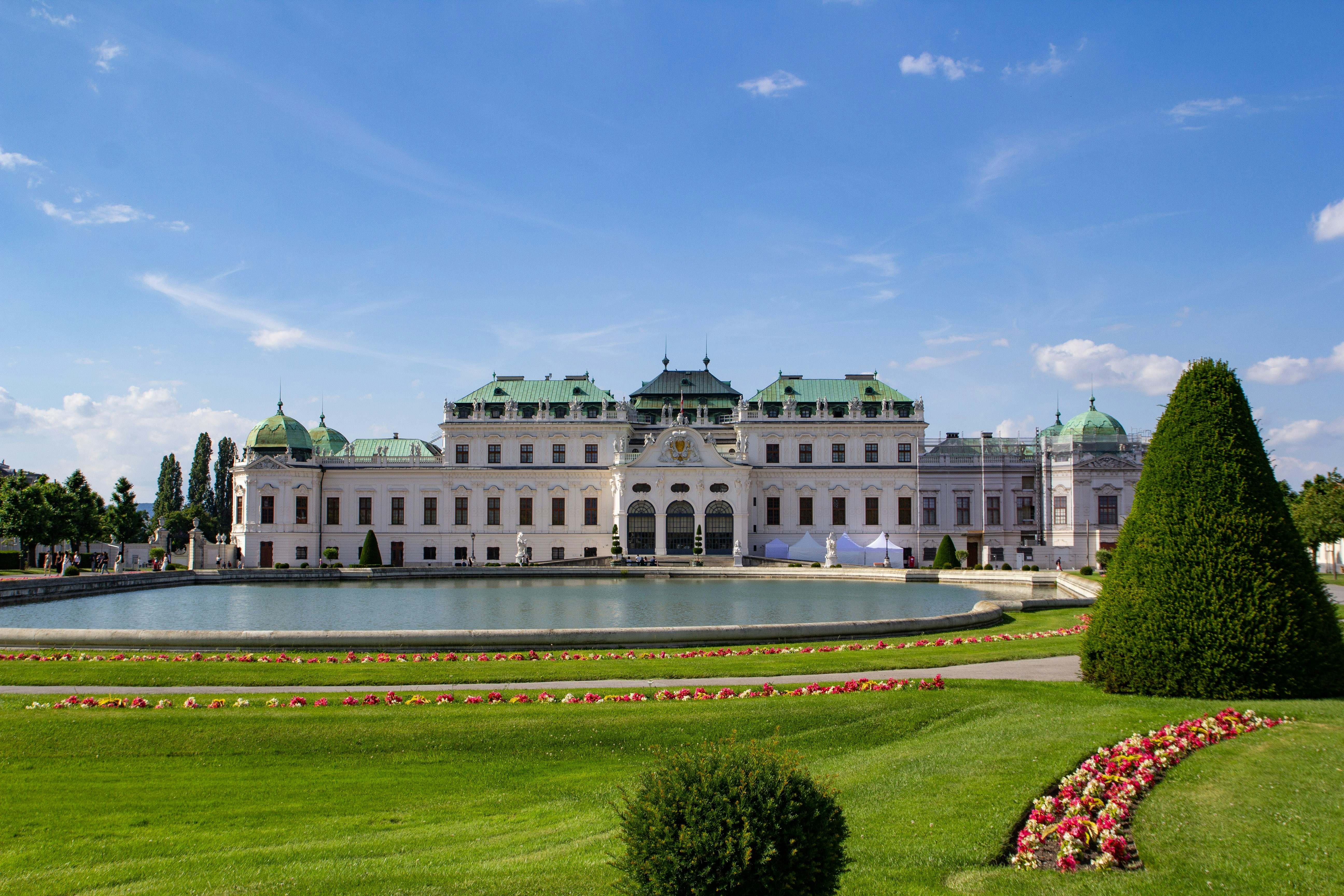 Elegant palace with green domes reflected in a tranquil canal, surrounded by manicured gardens and flowerbeds.