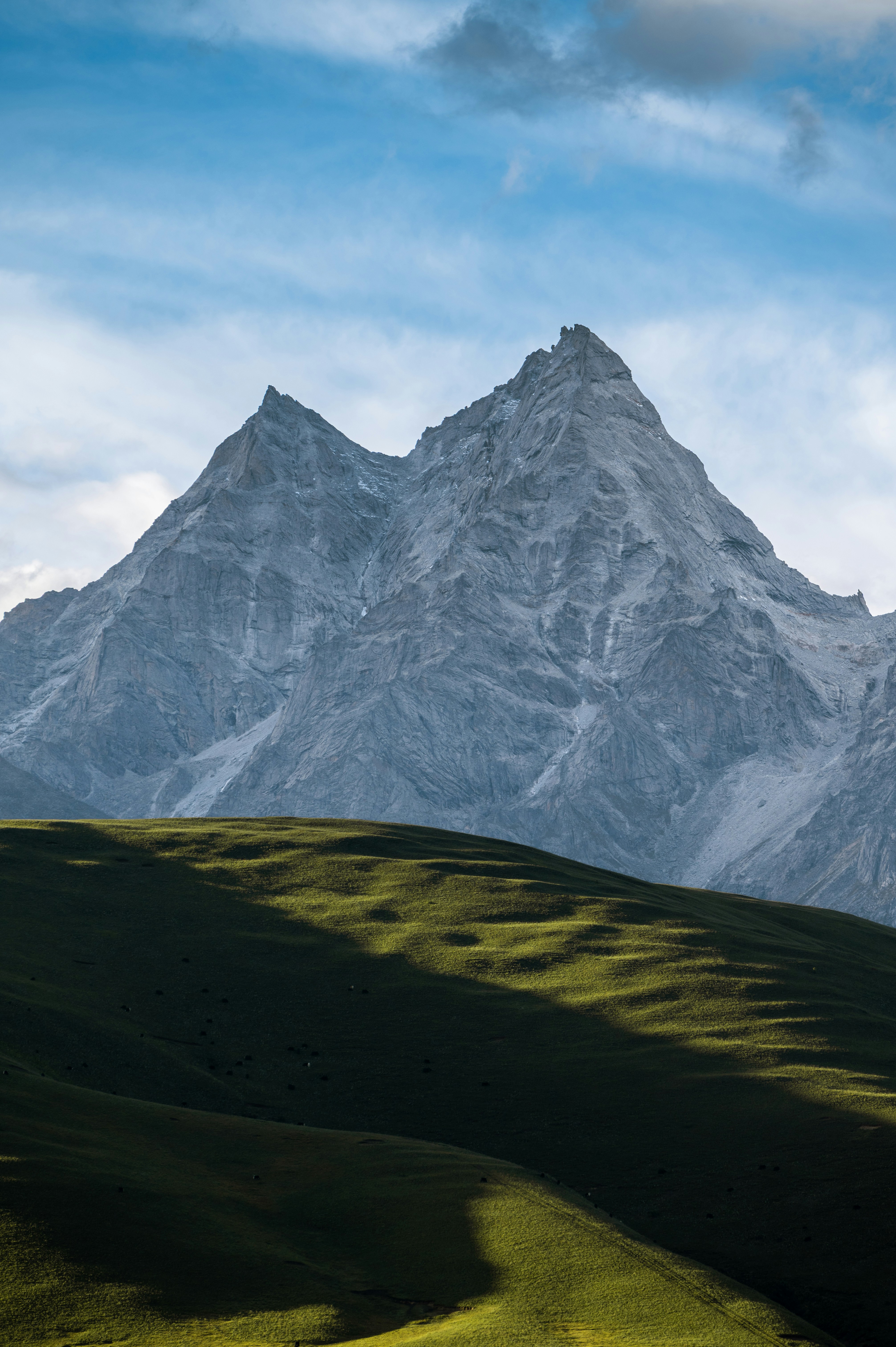 Rolling sunlit hills lead the eye toward jagged mountain peaks under a bright sky, capturing a serene alpine landscape.