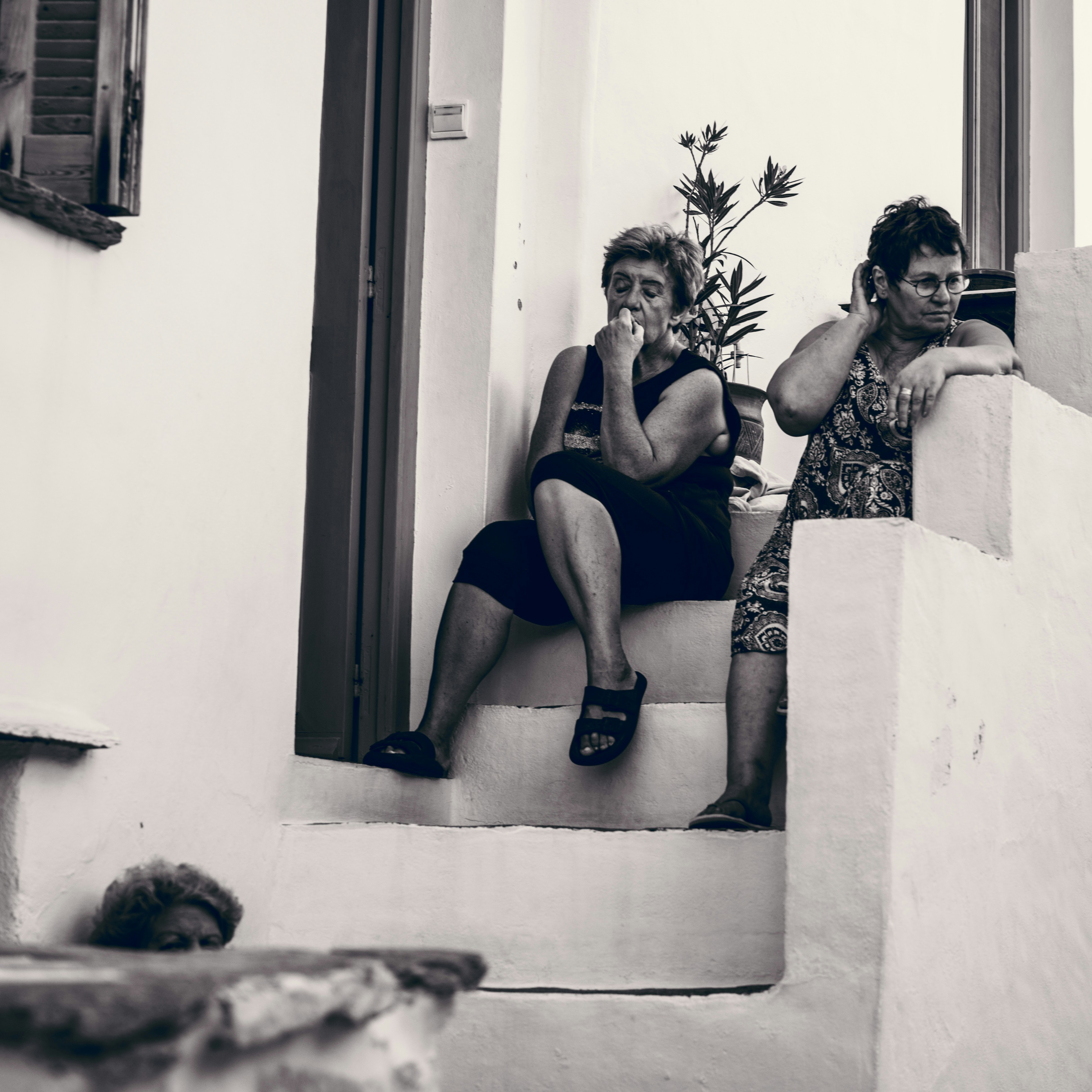 Two women sit on a sunlit white stair, one smoking and one talking on a phone, with a potted plant in the background; a partial head appears at the bottom left, all rendered in black and white.