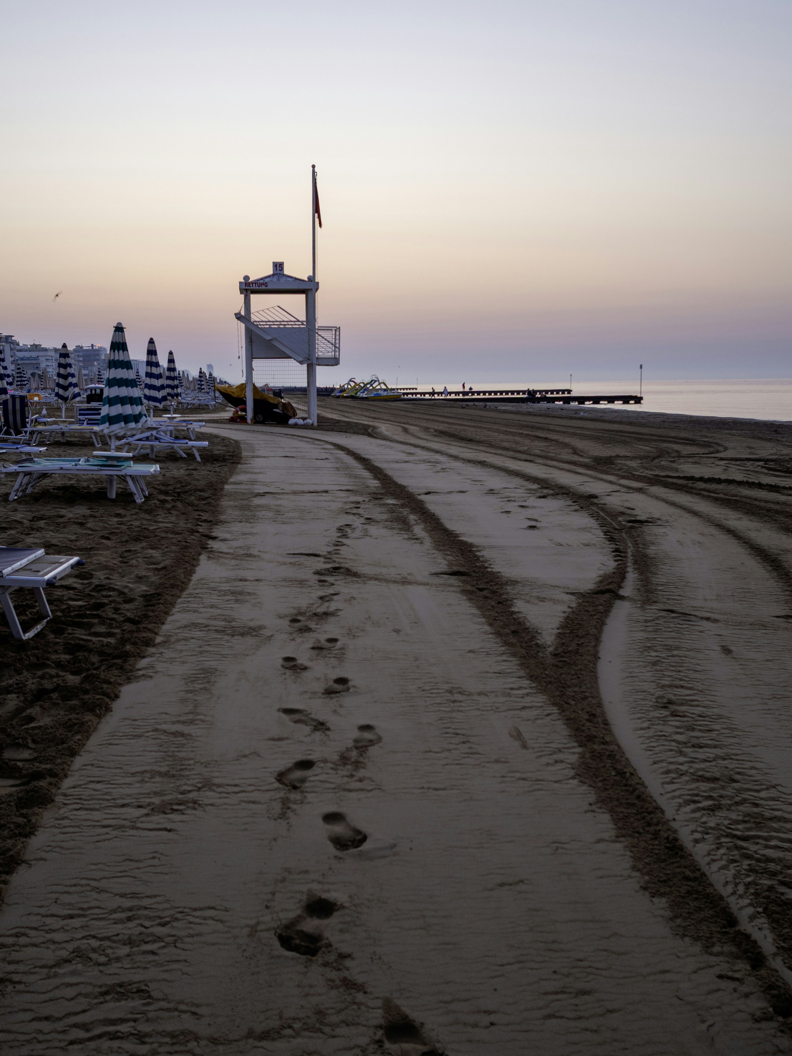 Sunlit beach with a lifeguard stand and empty lounge chairs along a sandy path leading to calm water at dawn. This is a photographic capture of a seaside resort scene with subtle color gradations and footprints in the wet sand.