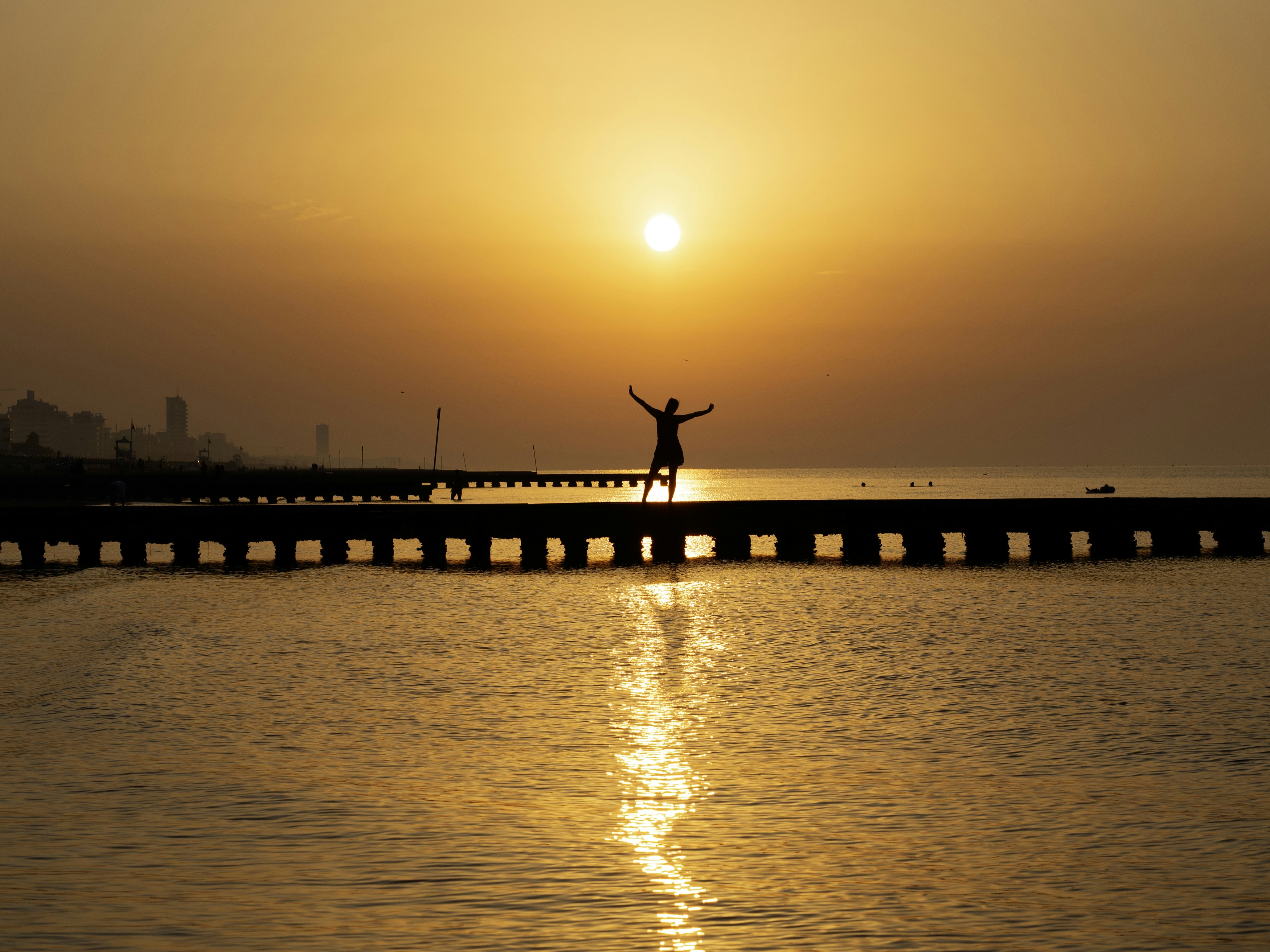 Silhouette of a person with arms raised balances on a sea-wacing pier at sunrise; warm orange sky and reflective water highlight the tranquil scene.