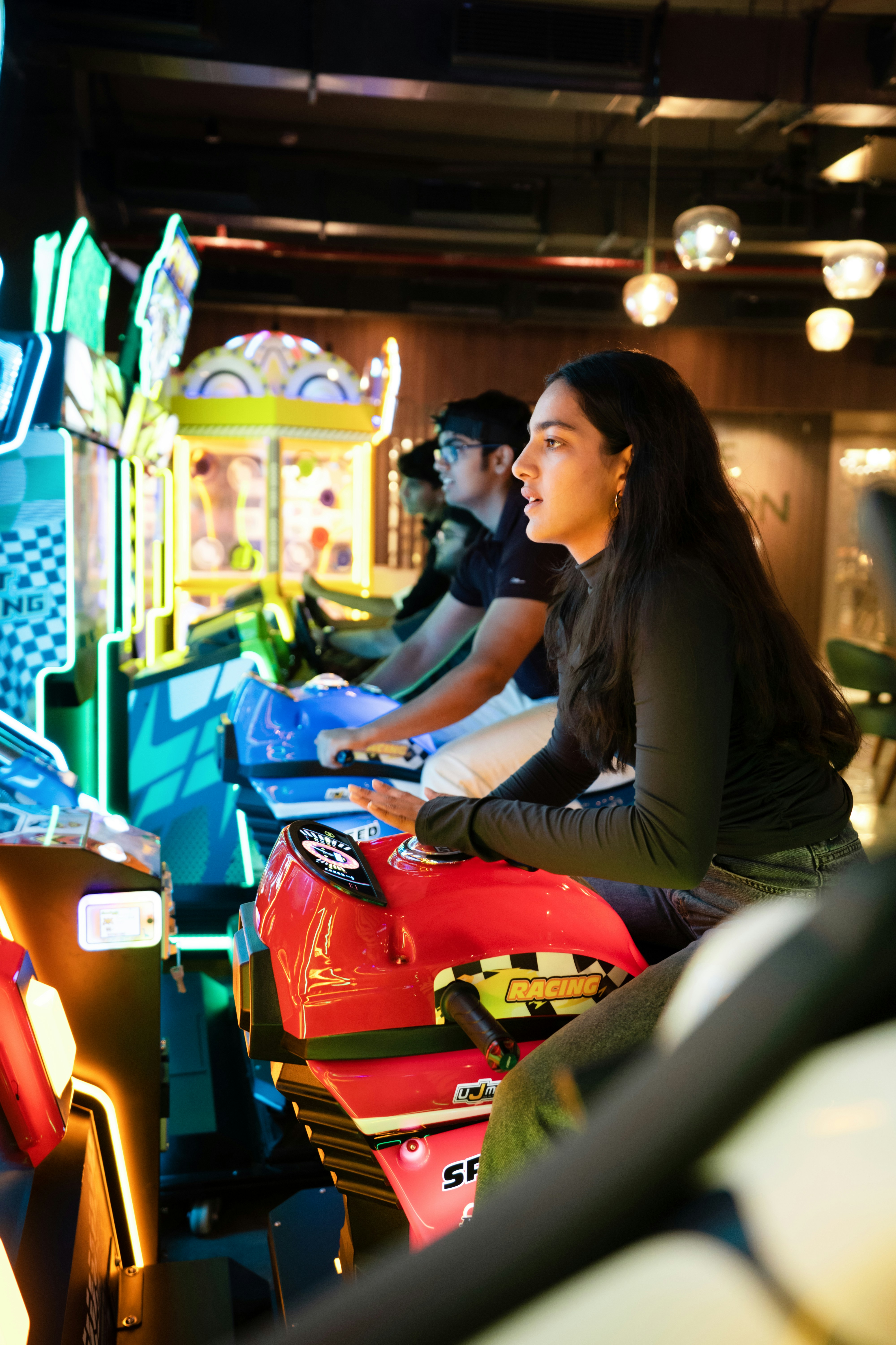 A young woman leans forward on a red arcade go-kart, with other players and glowing machines in the background, capturing a moment of concentration in an indoor game arcade.