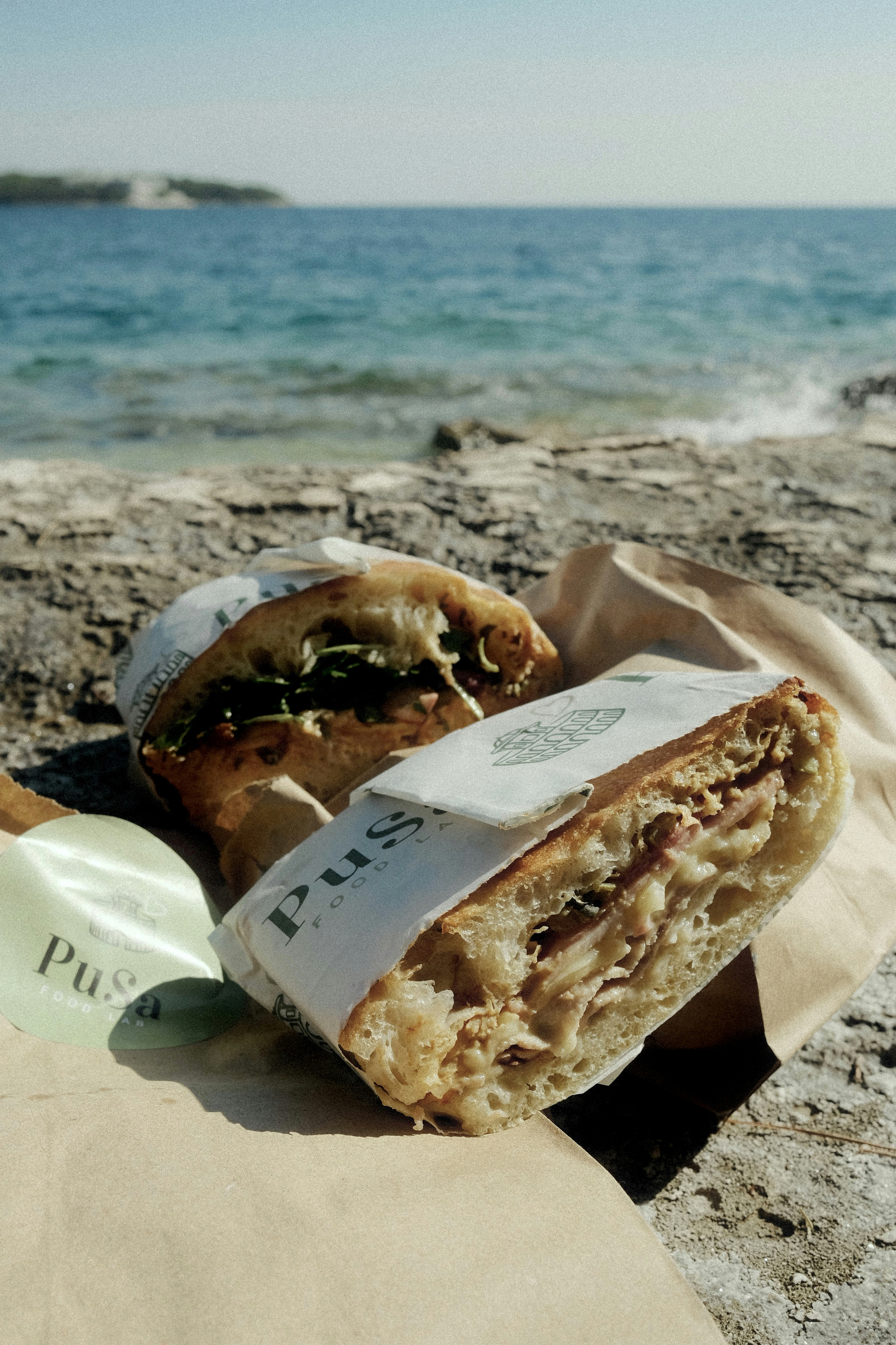 Sunlit seaside meal: a half-eaten pita sandwich rests on rocky shore