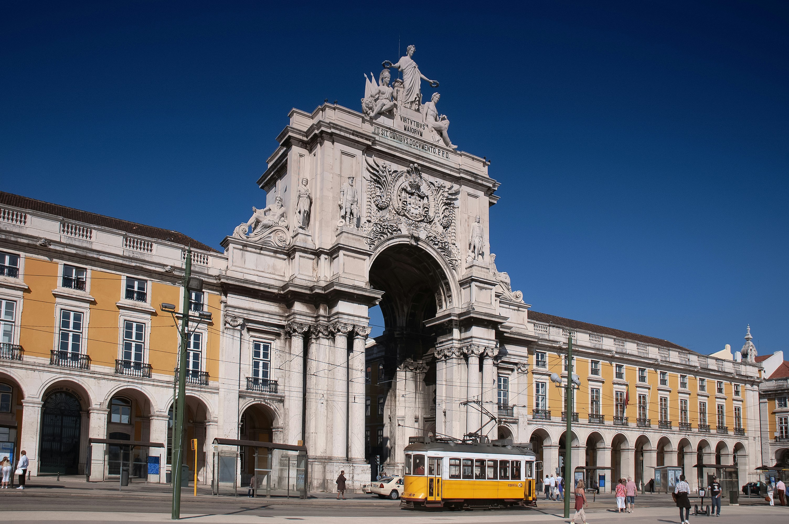 Historic yellow electric tram and streetcar driving past the monumental Rua Augusta triumphal arch and yellow arcade buildings at Praca do Comercio tourist destination, Lisbon, Portugal.