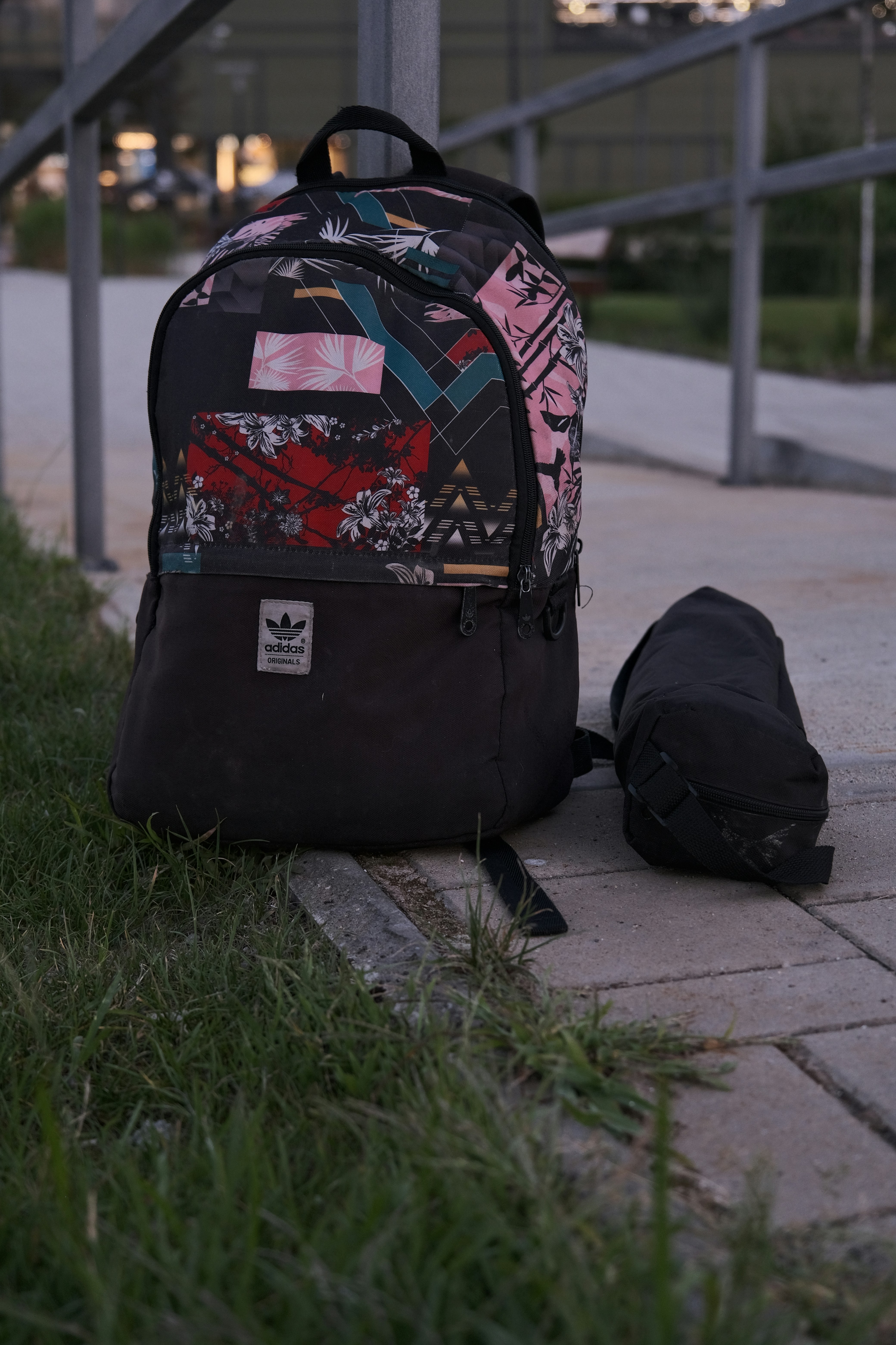 Colorful patterned backpack sits upright on a concrete path with a compact bag to its right; grassy edge and urban railing in background. This is a lifestyle product shot focused on the backpack design.