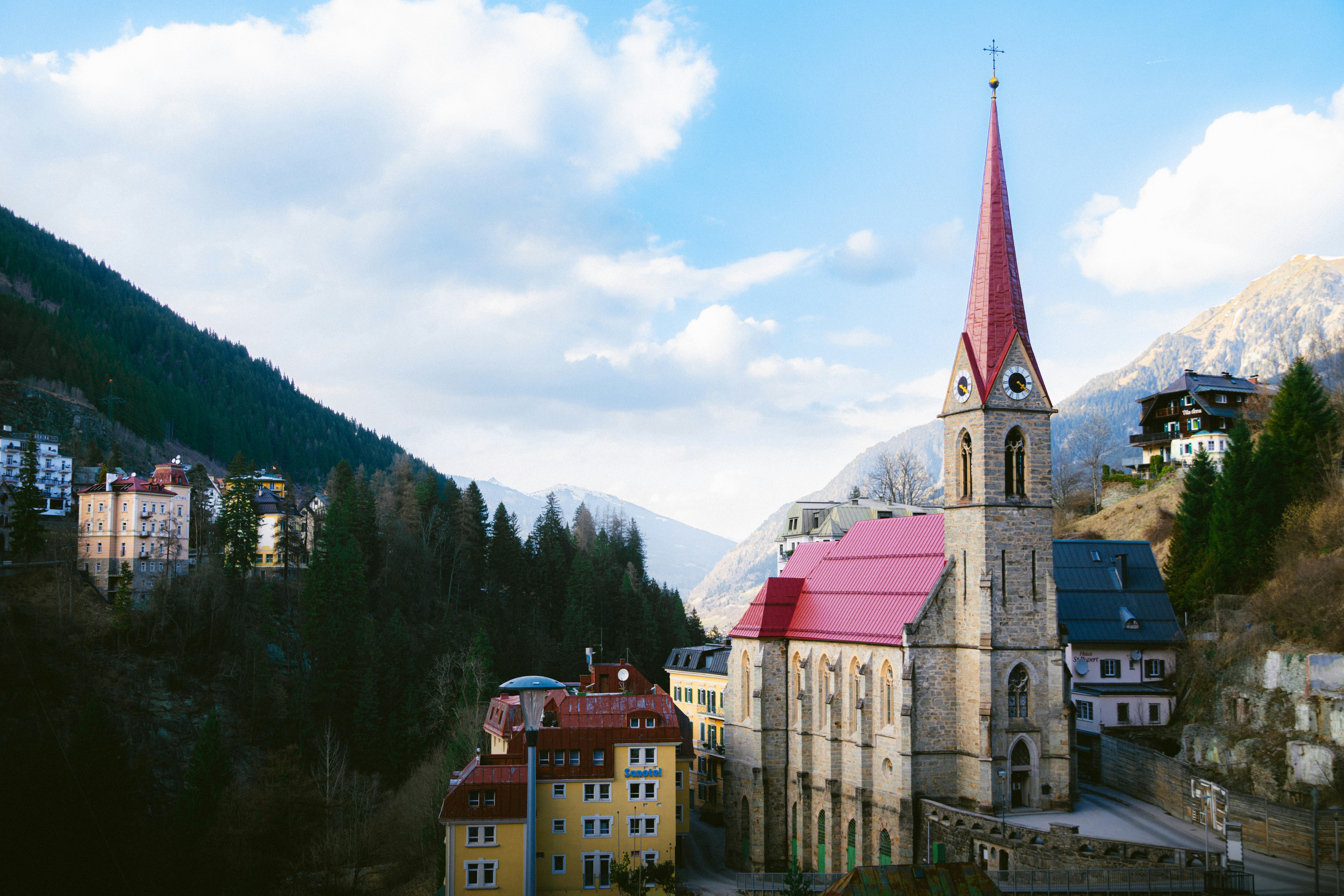 Mountain village with a prominent church and red roof spire amid colorful houses, set against forested hills and snow-capped peaks.