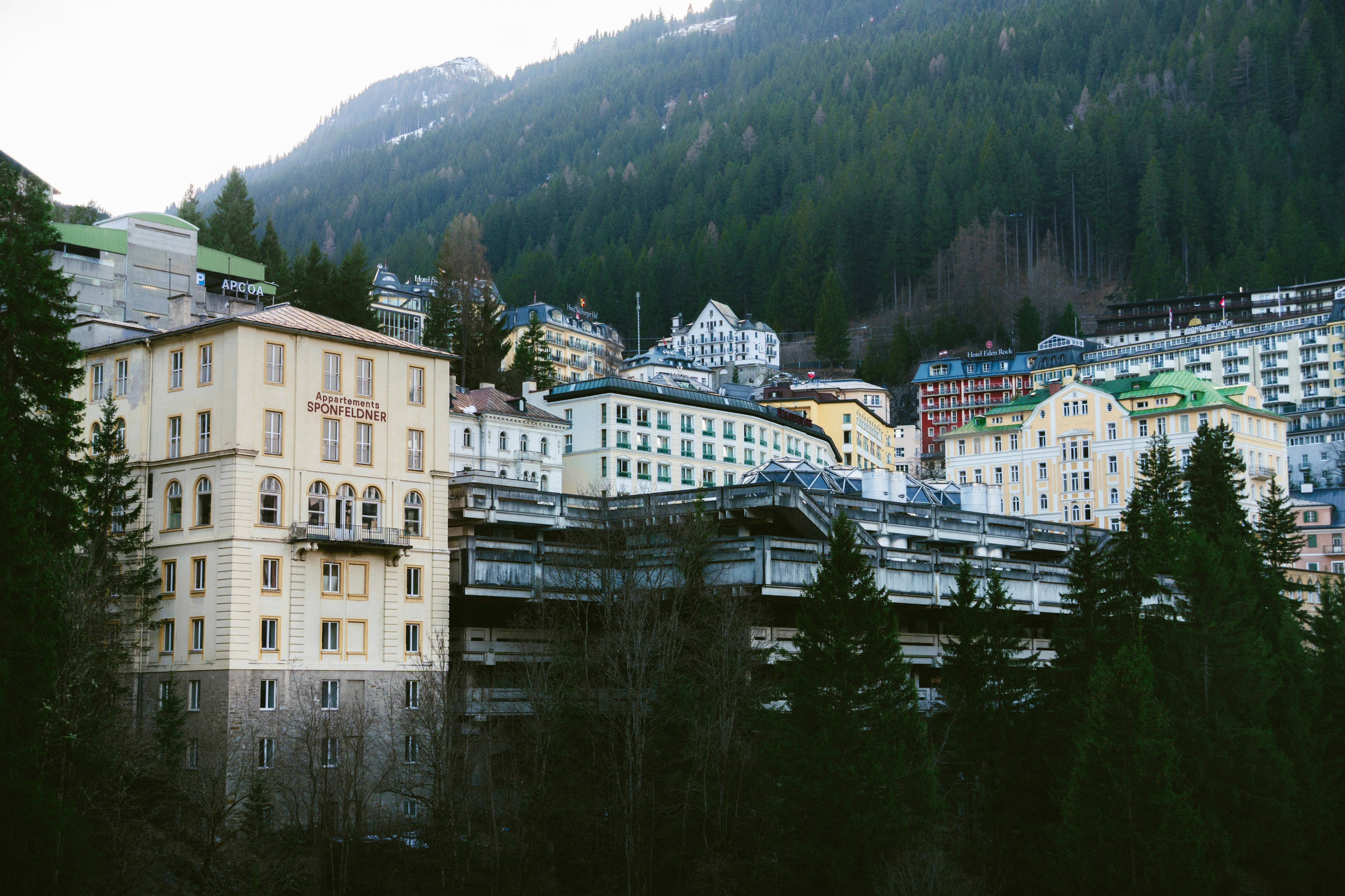 Colorful hillside buildings cascade up a forested slope, with a mix of pastel facades and green roofs forming a layered cityscape.