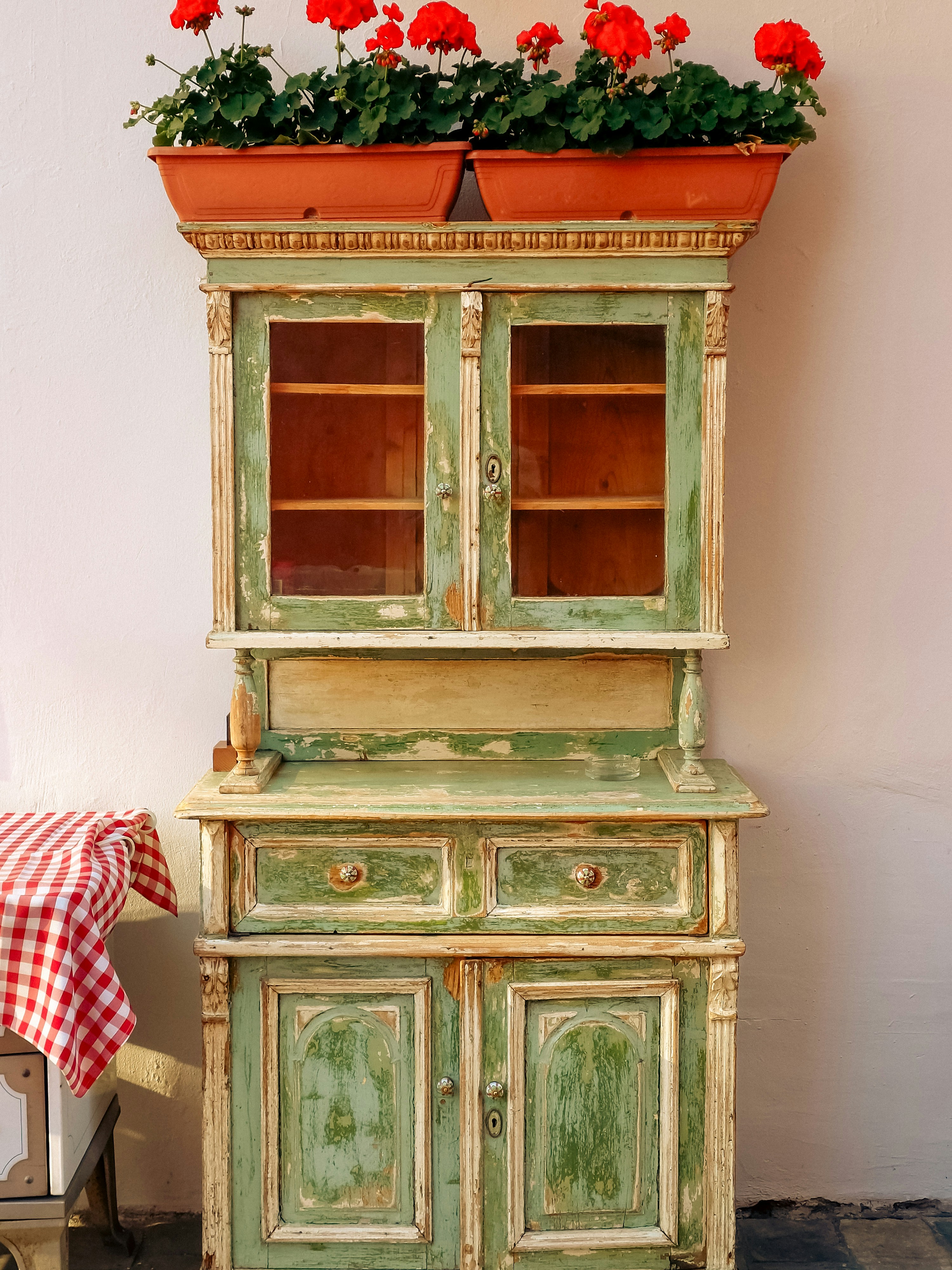 Weathered green cabinet with glass doors and lower cupboards sits against a pale wall; a planter with red geraniums rests atop, and a checkered tablecloth peeks from the left. A rustic, vintage furniture vignette.