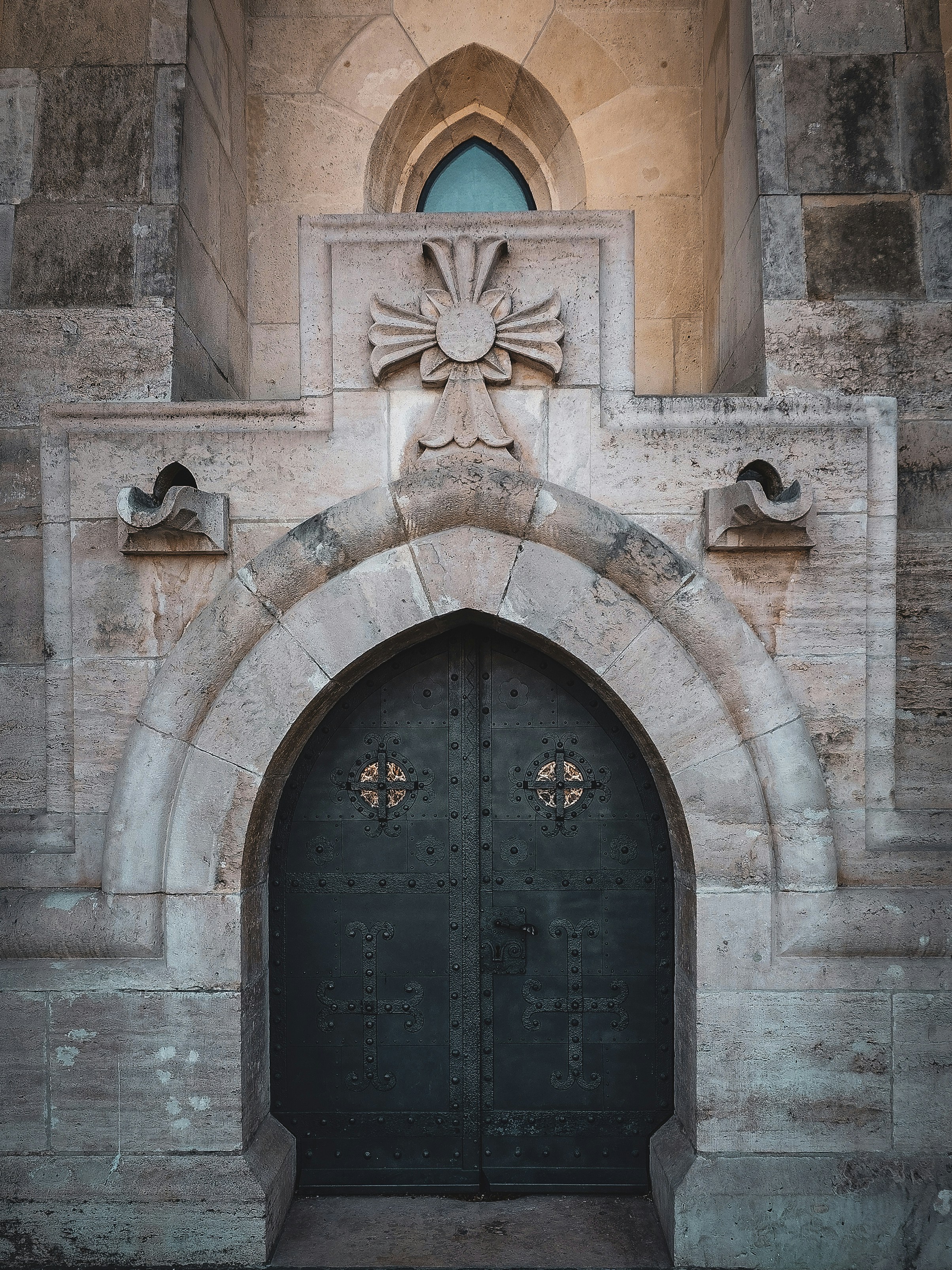 Dark iron double doors set in a carved stone arch with a cross motif above, part of a weathered stone church facade.