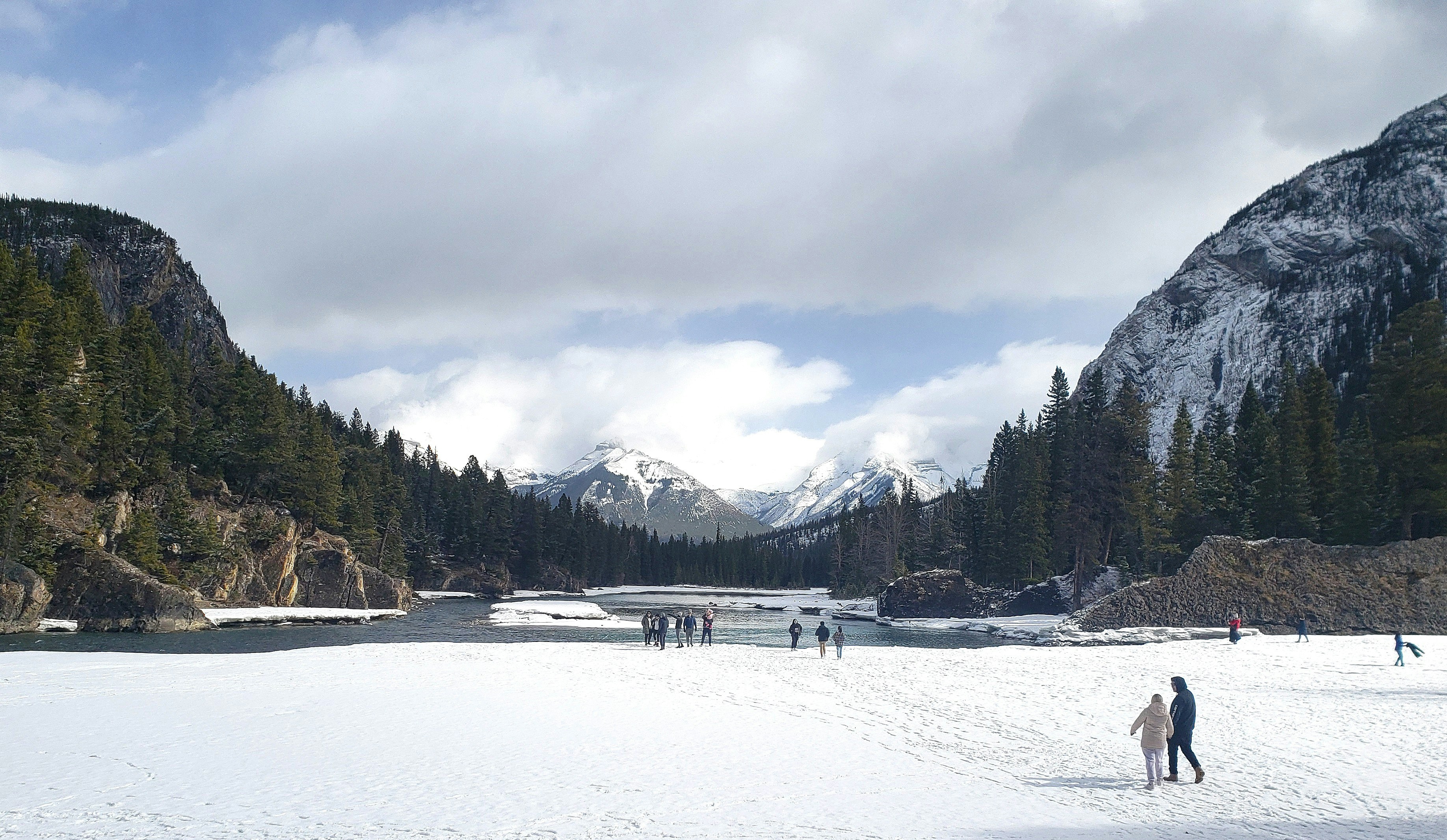Expansive snowy valley framed by tree-dotted mountains under a cloudy sky, with hikers and a couple strolling on a frozen river. A tranquil alpine scene highlighting landscape scale.