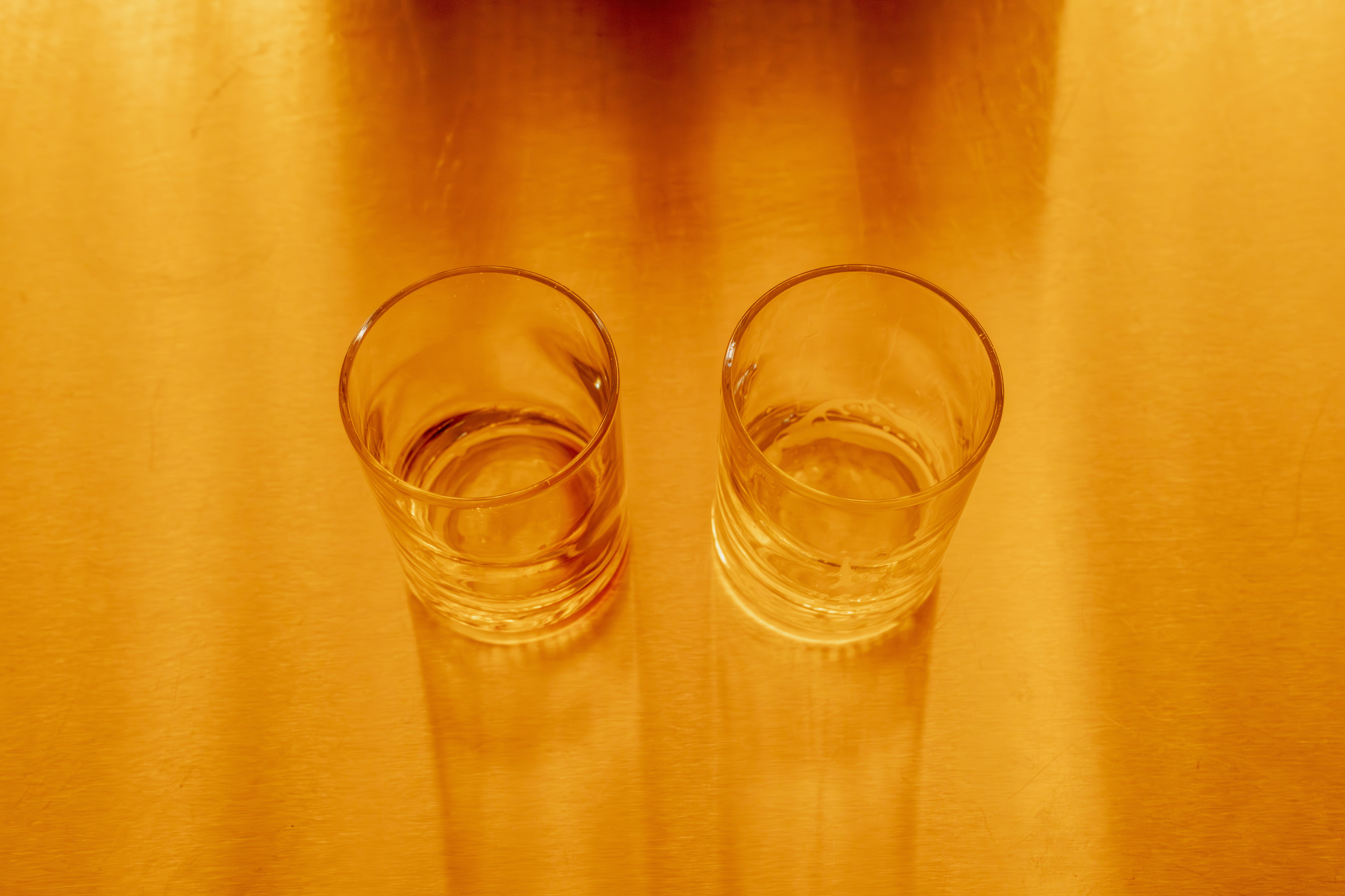 Two transparent glasses sit on a warm amber-toned surface, casting soft reflections and subtle shadows in a close-up still life.