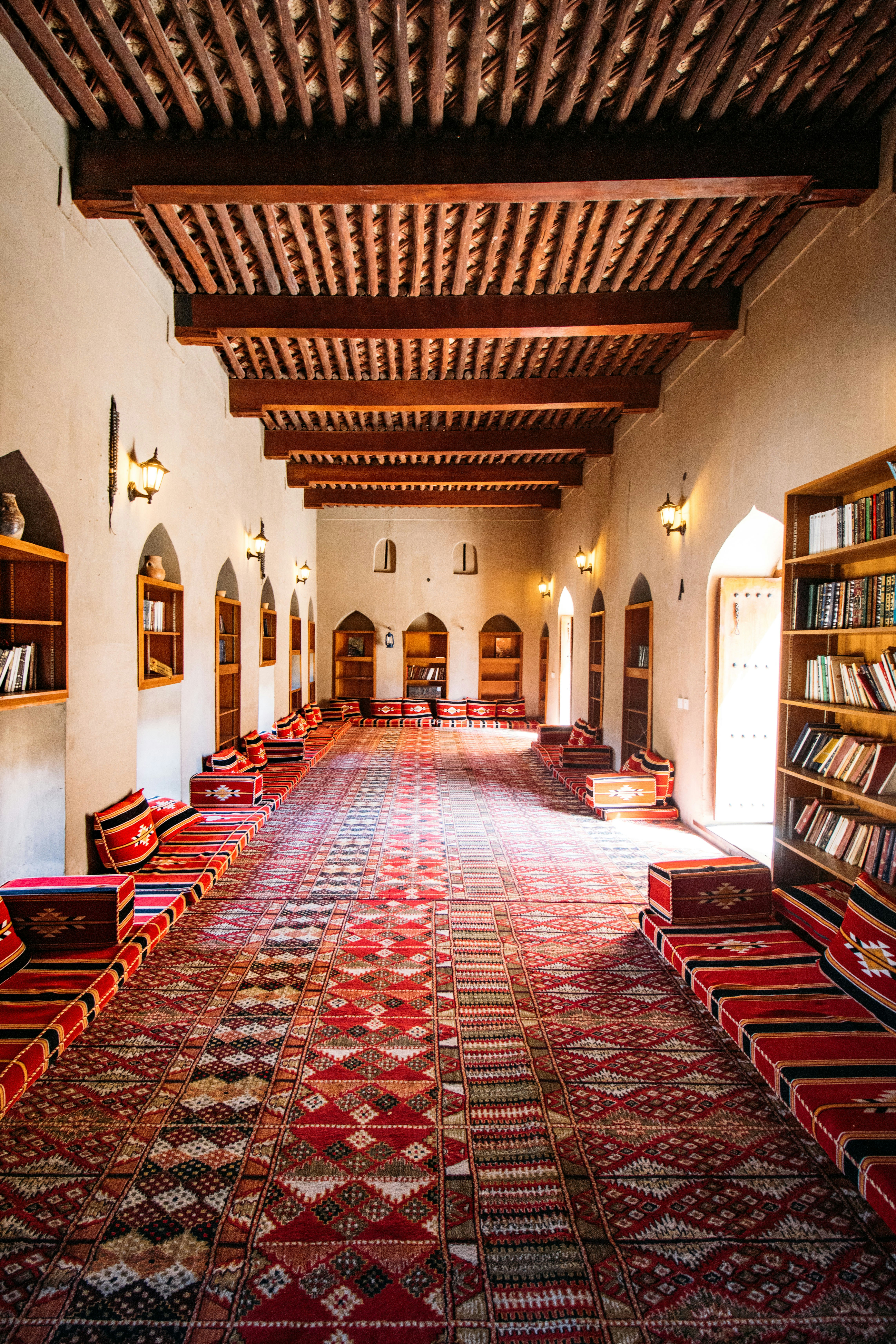 Long library hall with warm lighting, carved wooden ceiling beams, archways, and low cushioned benches along patterned carpet.