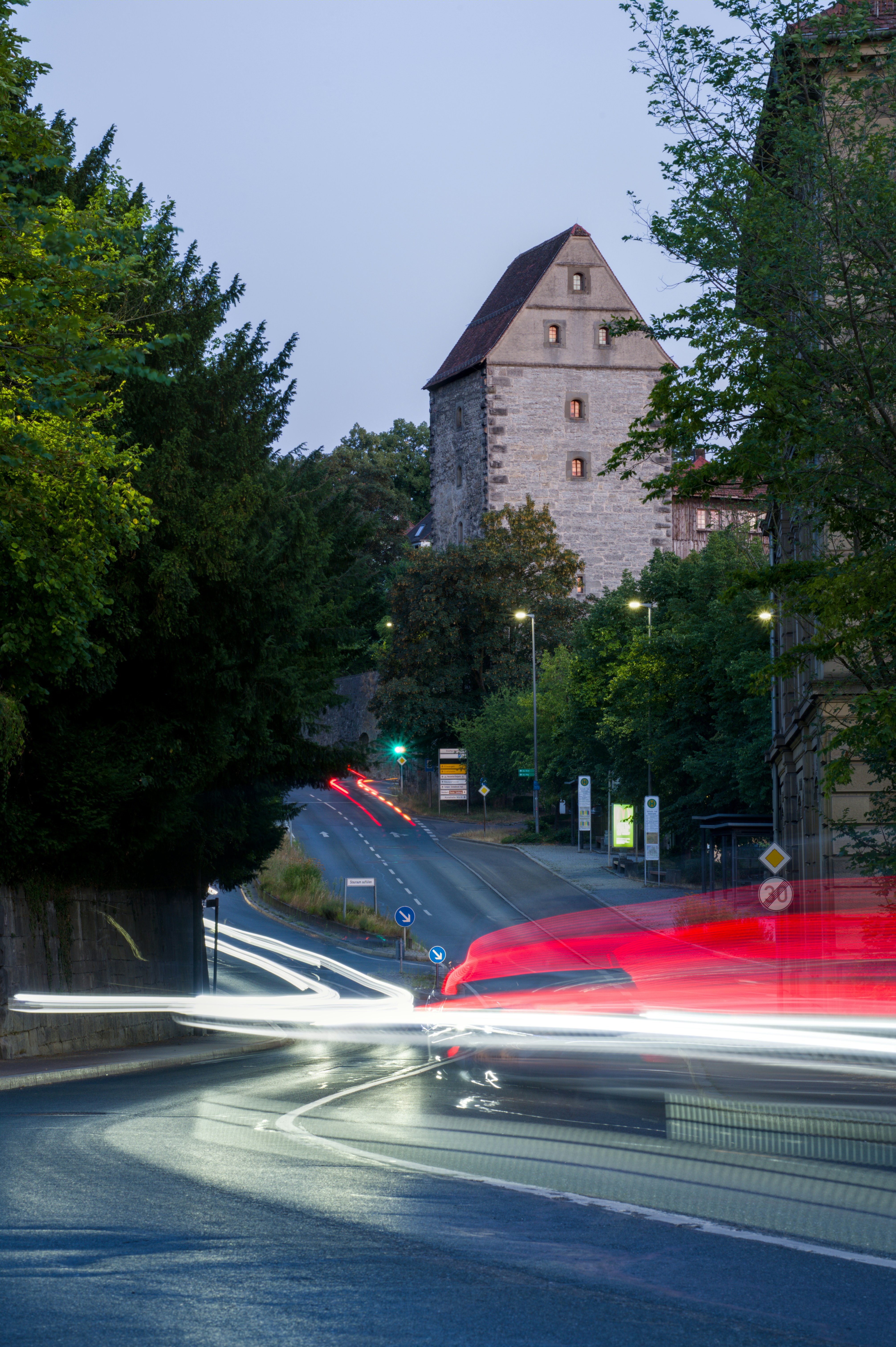 Stone tower rising beyond a tree-lined street as car taillights streak across a waning blue hour; the scene blends ancient architecture with modern traffic in a quiet town slope.