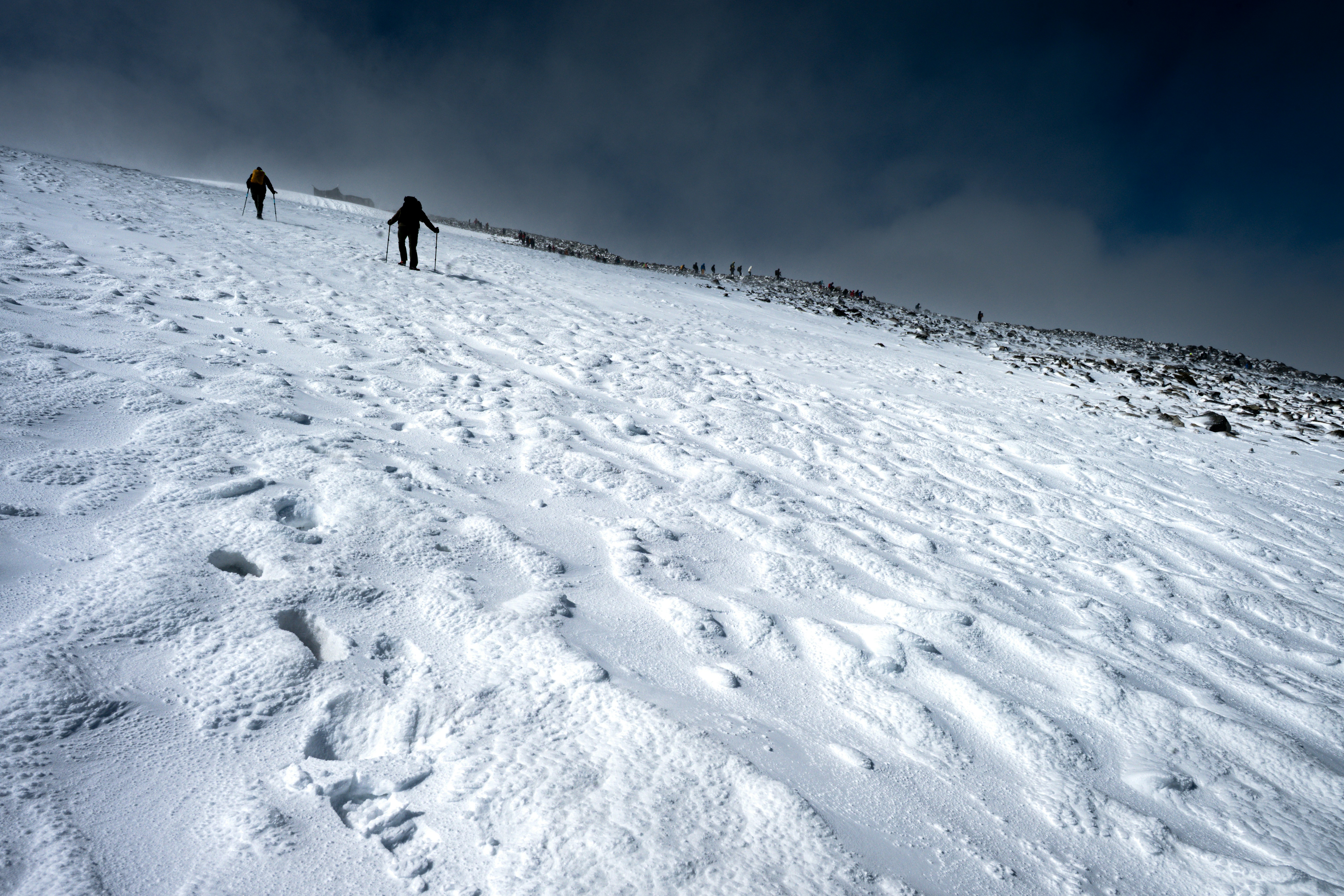 Hikers cross a snow-covered slope on the ascent from Spiterstulen to Galdhøpiggen, the highest mountain in Norway, rising 2,469 metres above sea level. The demanding trail leads through the high-alpine landscape of Jotunheimen, where snowfields, exposed terrain and rapidly changing weather shape the journey to the summit. Part of “The Crown of Europe” series documenting ascents to the highest peaks of European countries.