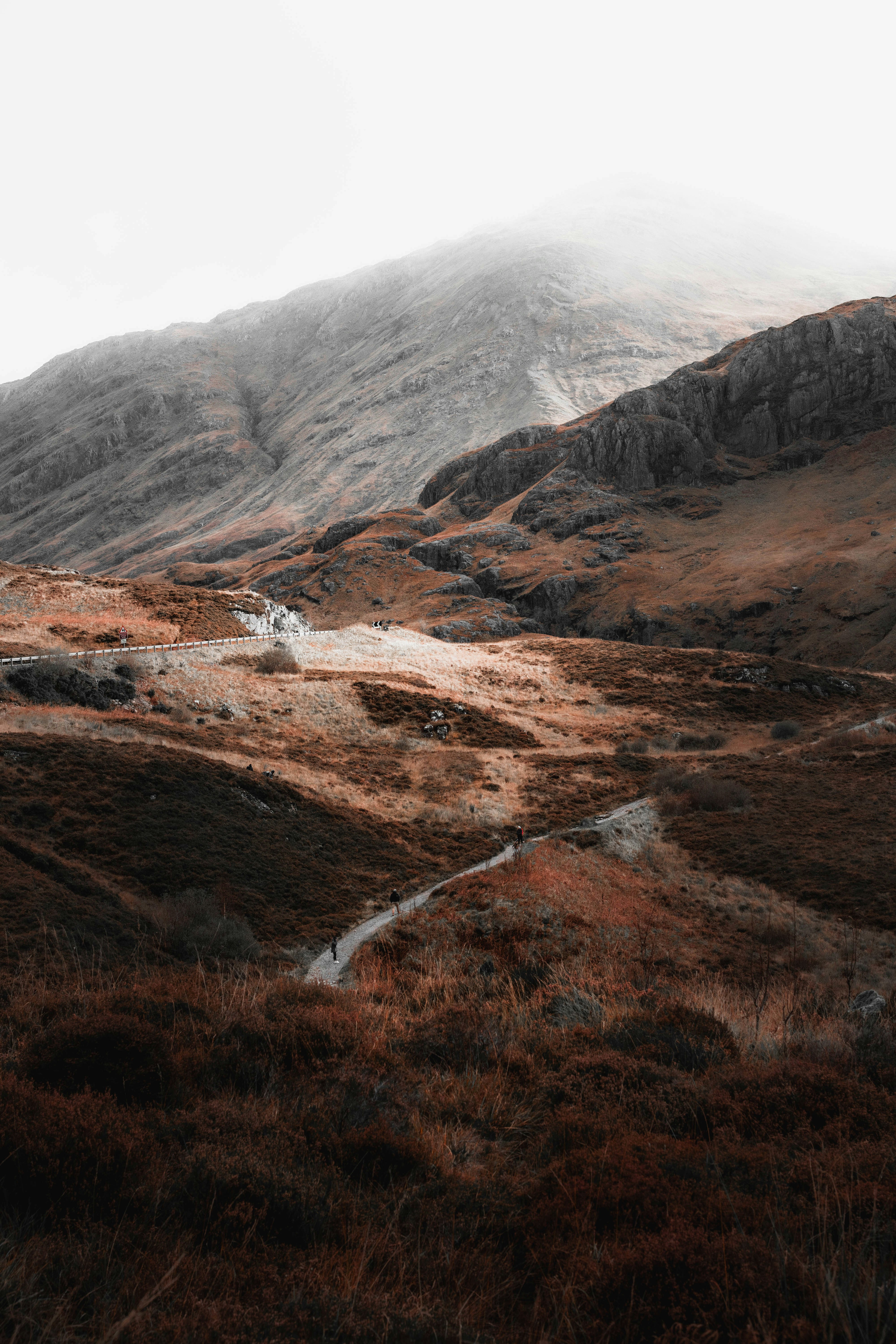 Dusty trail winds through rugged hills and heather-dotted moorland toward a distant misty peak.