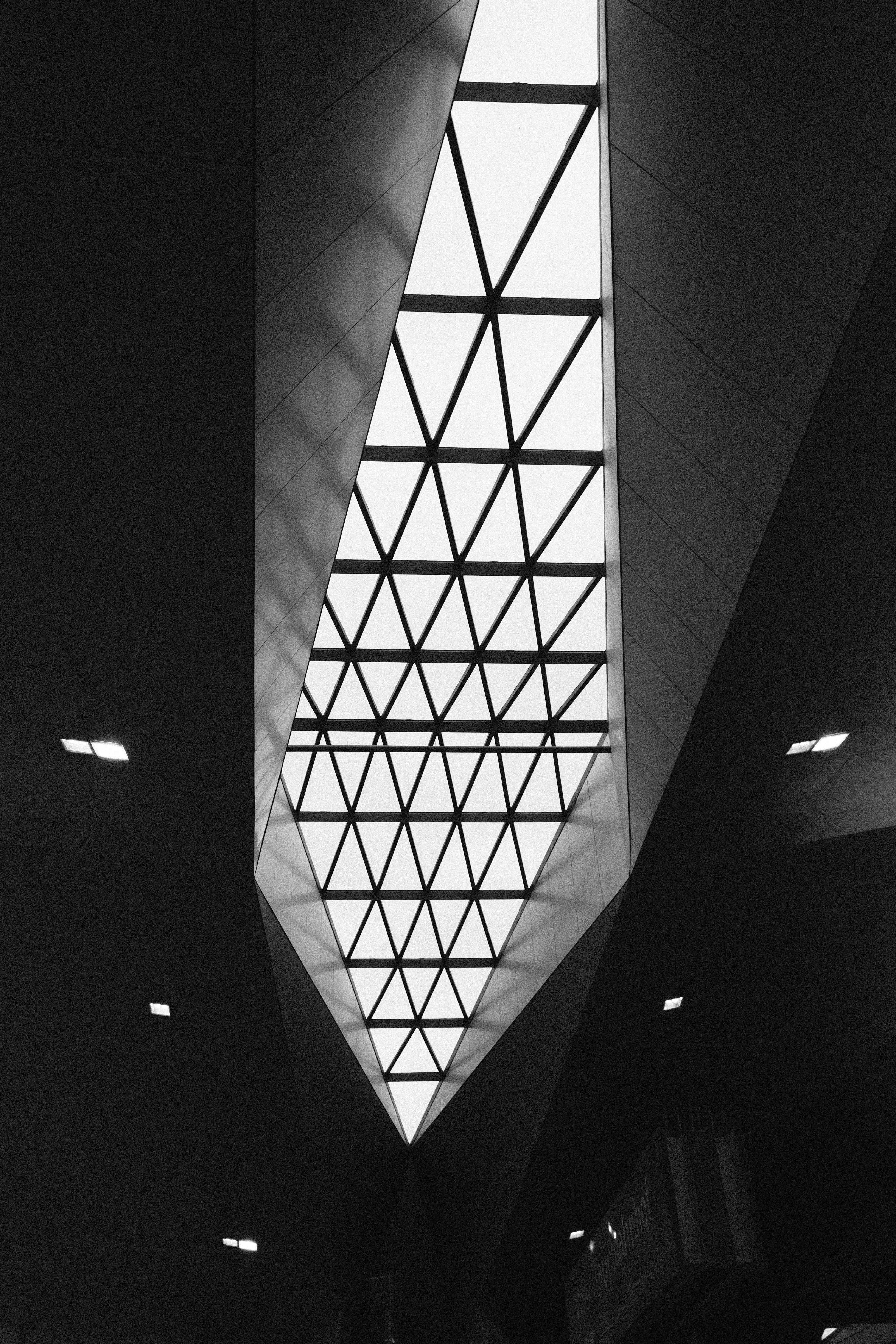 A stark black-and-white interior shot focusing on a triangular glass roof lattice framed by dark architectural walls.