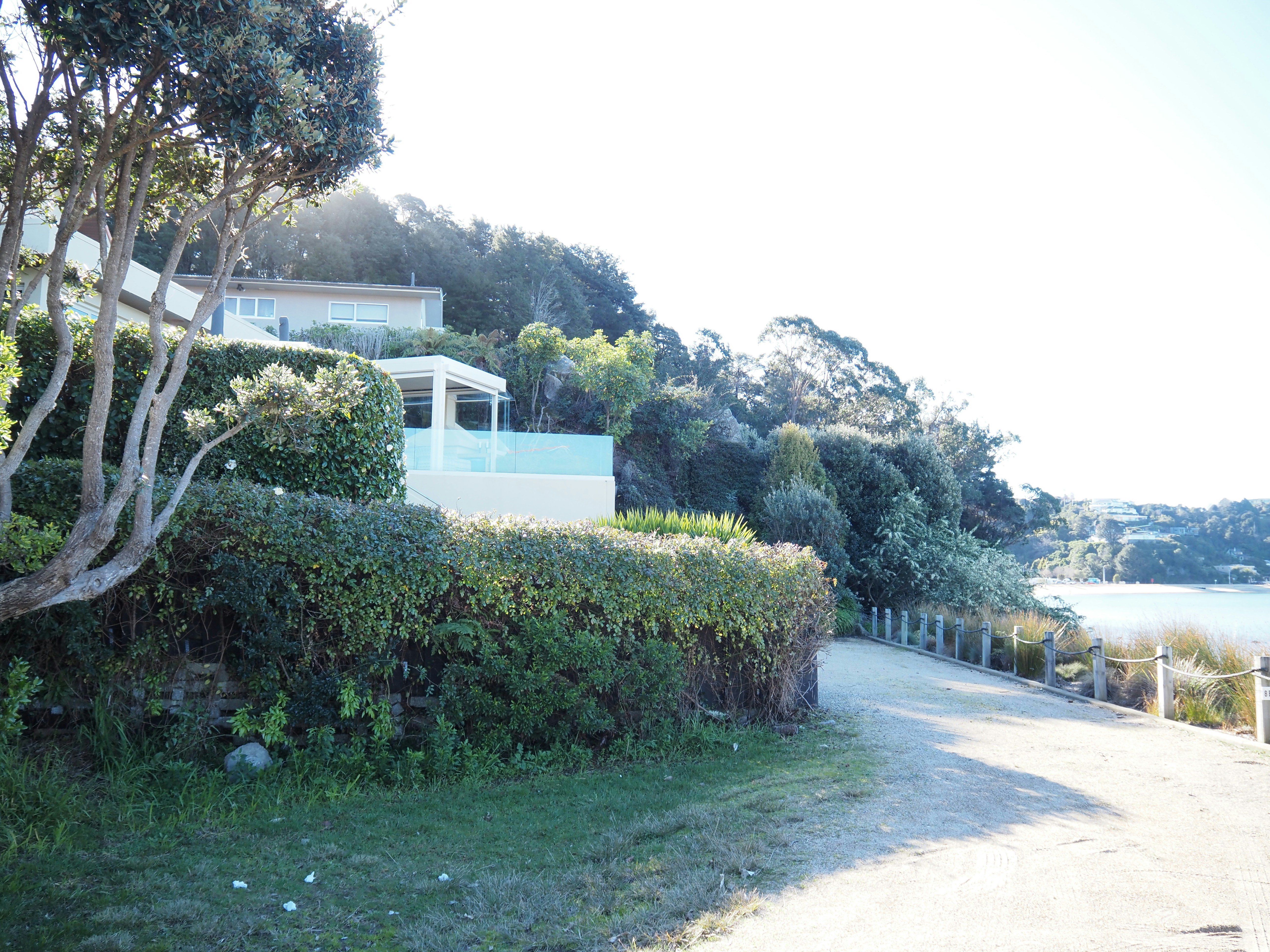 Sunlit coastal path lined with trimmed hedges and a modern house to the left, leading toward the water on the right; the scene is bright with minimal clouds and natural greenery.