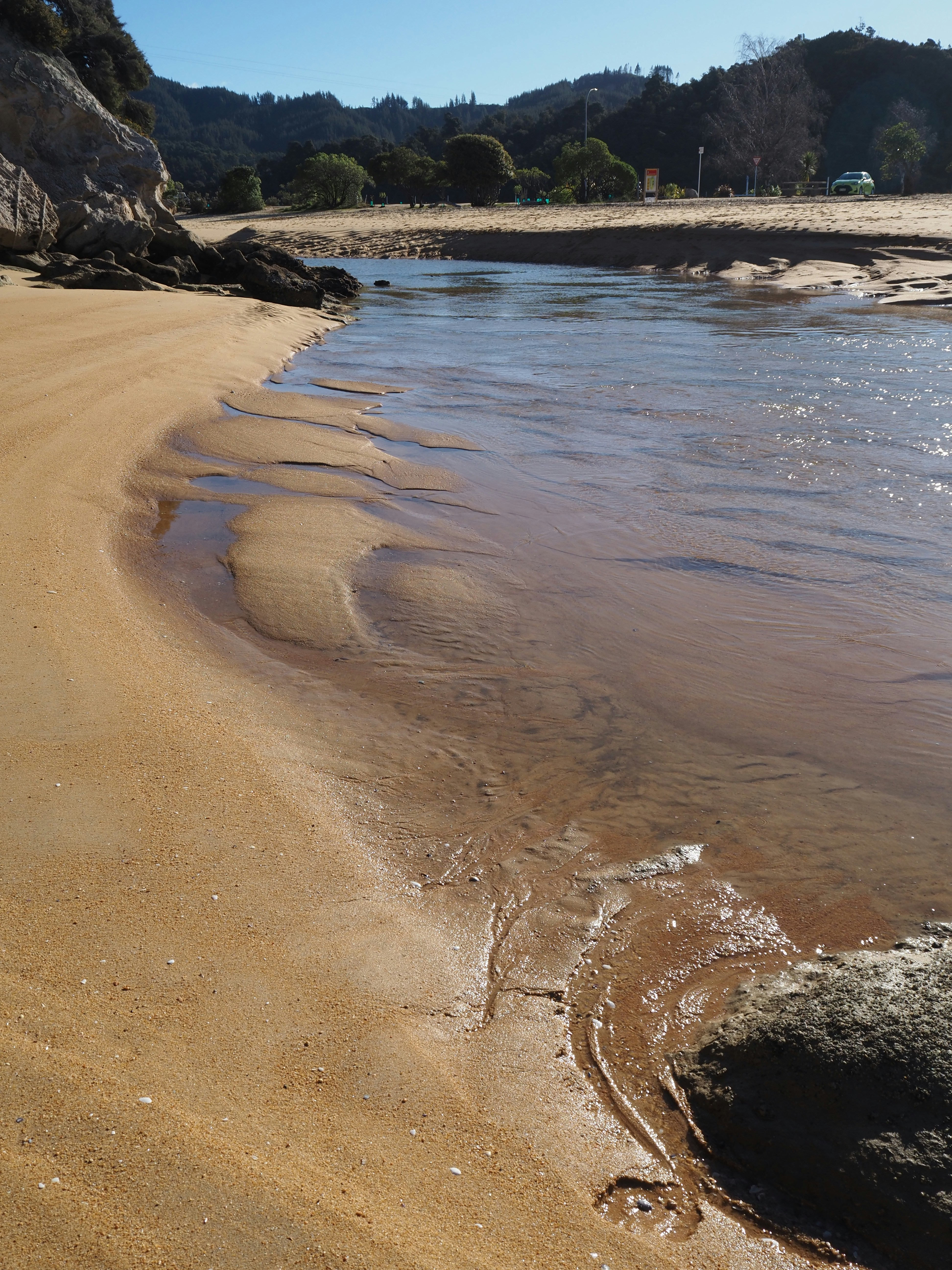 Sunlit shoreline with receding tide carving ripples in wet sand along a calm inlet; distant hills and trees form a quiet backdrop at the top of the frame. This is a natural landscape capture.