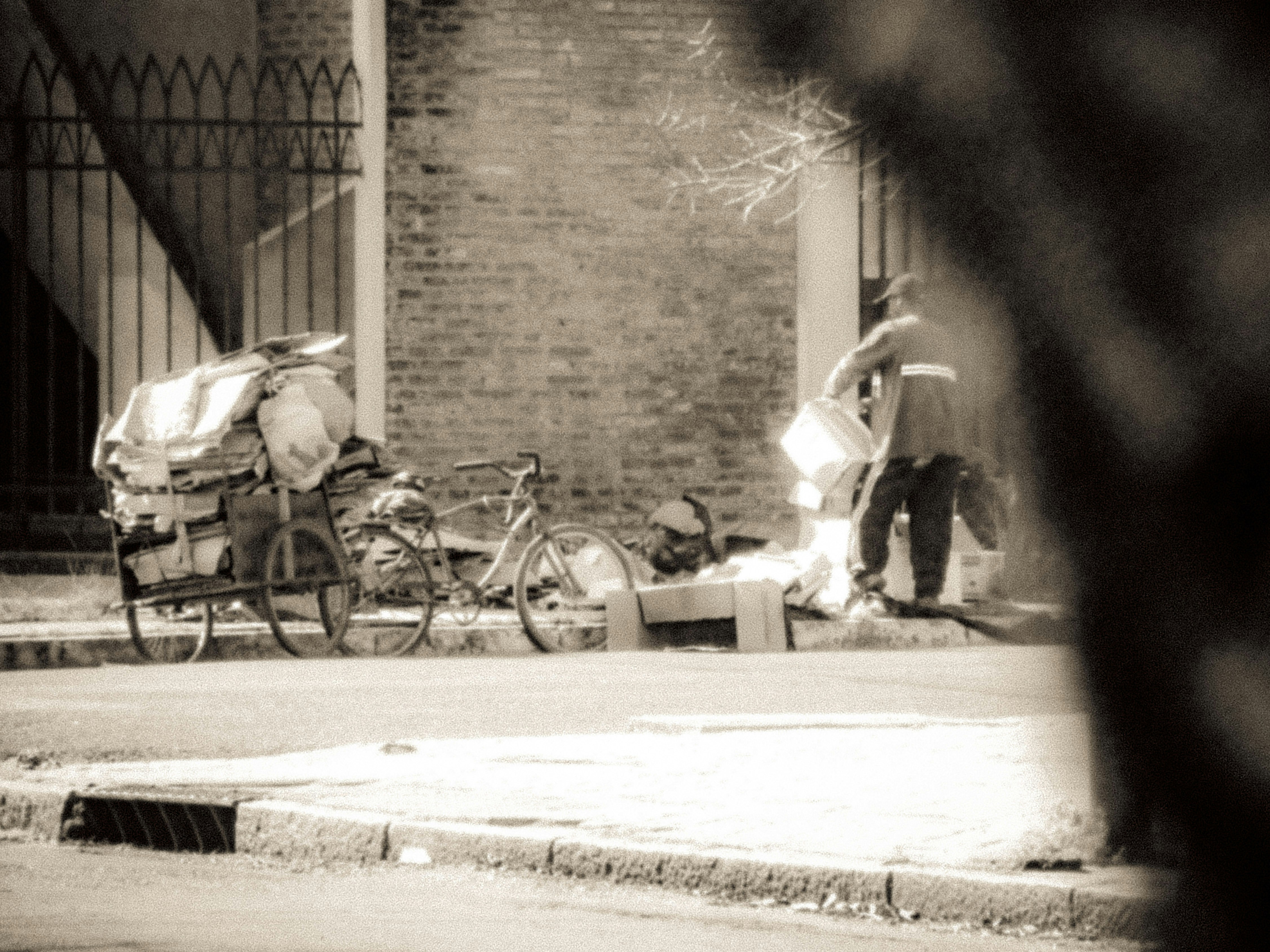 Street scene with a bicycle cart and a person arranging items on a curb, backlit by warm, sepia-toned light; includes a logbook of boxes and a distant figure near a doorway, suggesting daily urban activity.