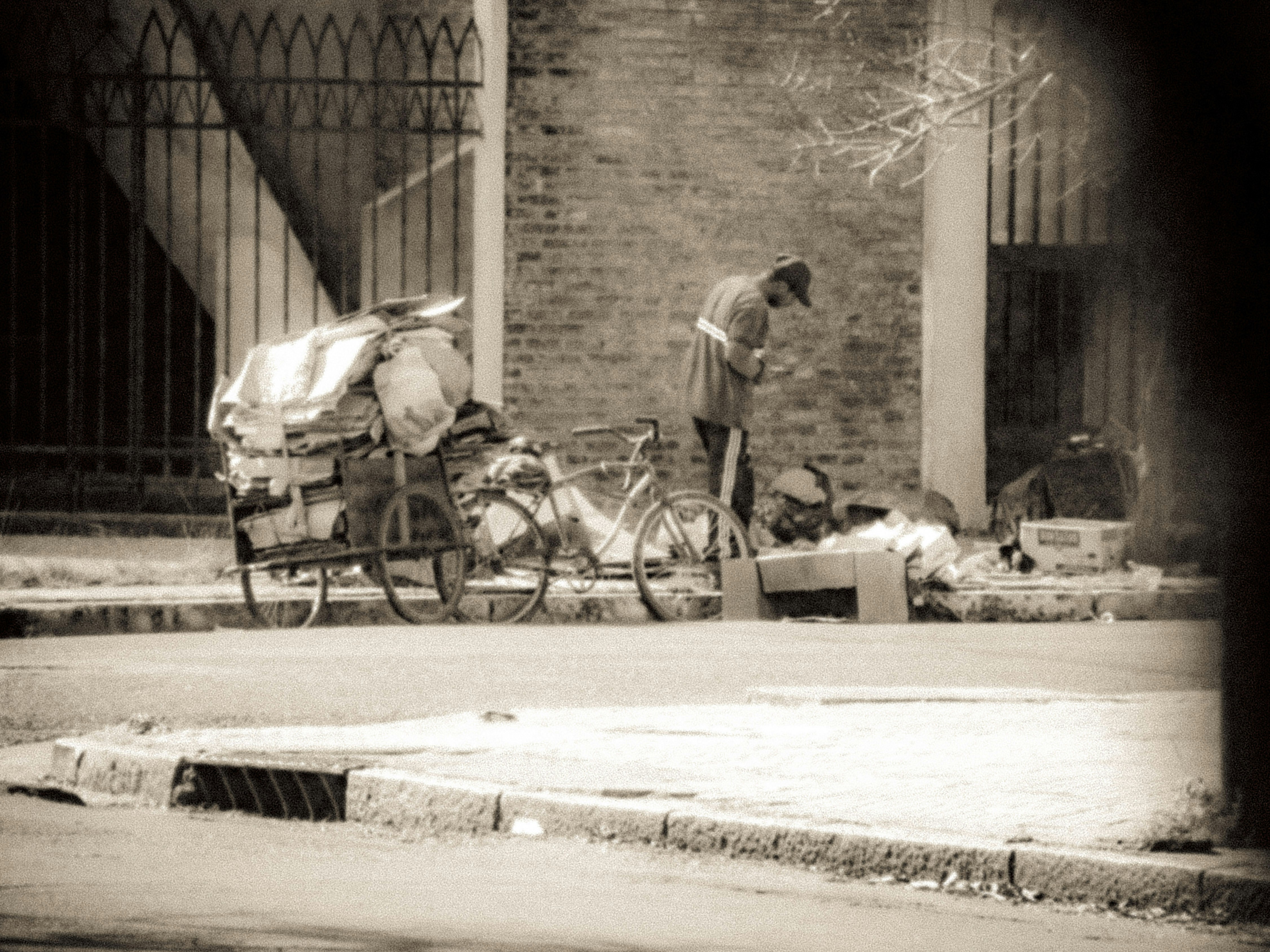 Two bicycles and a person rummaging through a pile of debris on a quiet sidewalk; monochrome street scene with a brick wall and iron fence in the background.