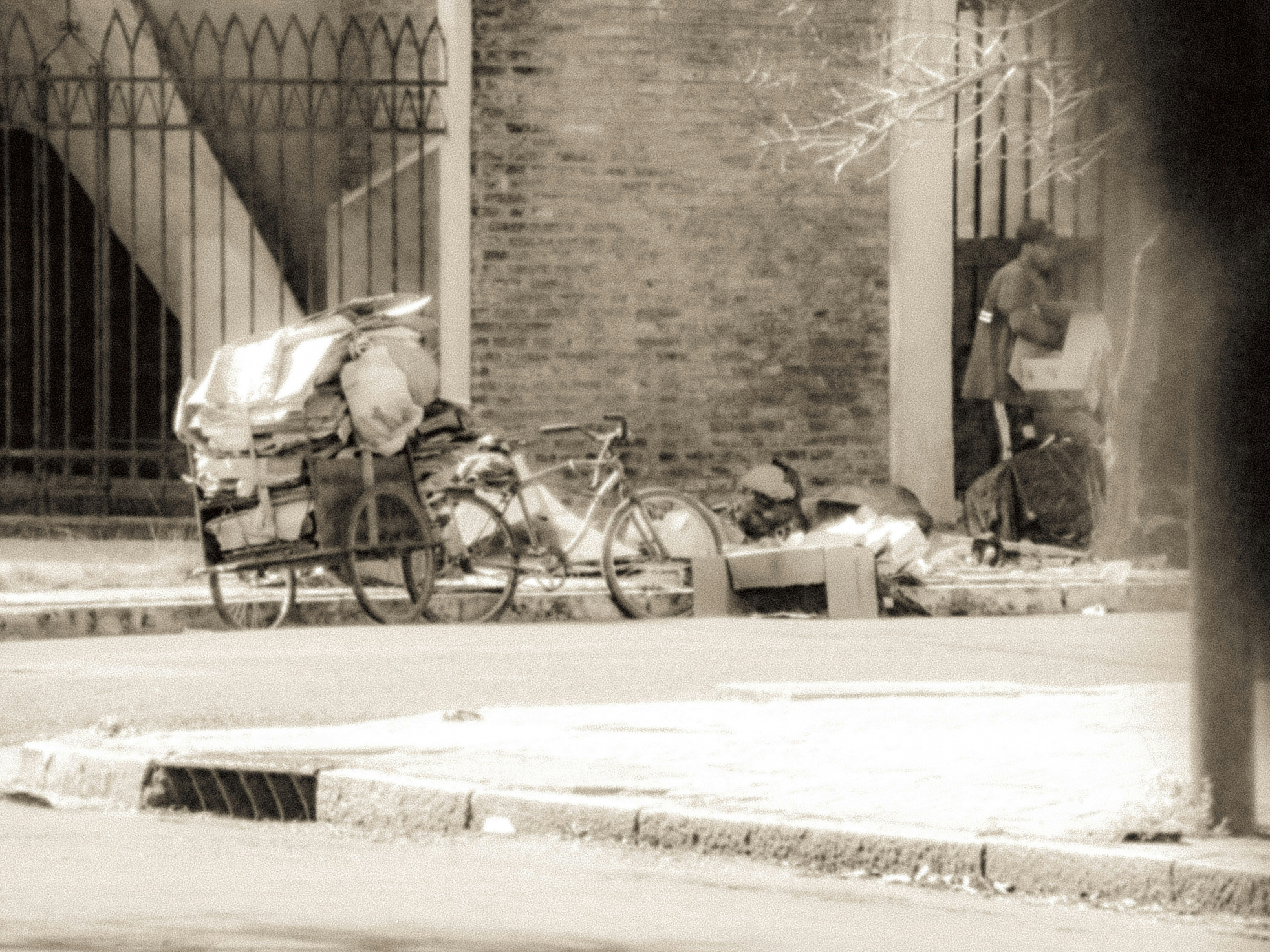 Street scene with a loaded bicycle rickshaw-style cart and scattered items by a curb; a lone figure in the background repositories a box near a brick wall. A muted, sepia-toned vibe emphasizes urban stillness.