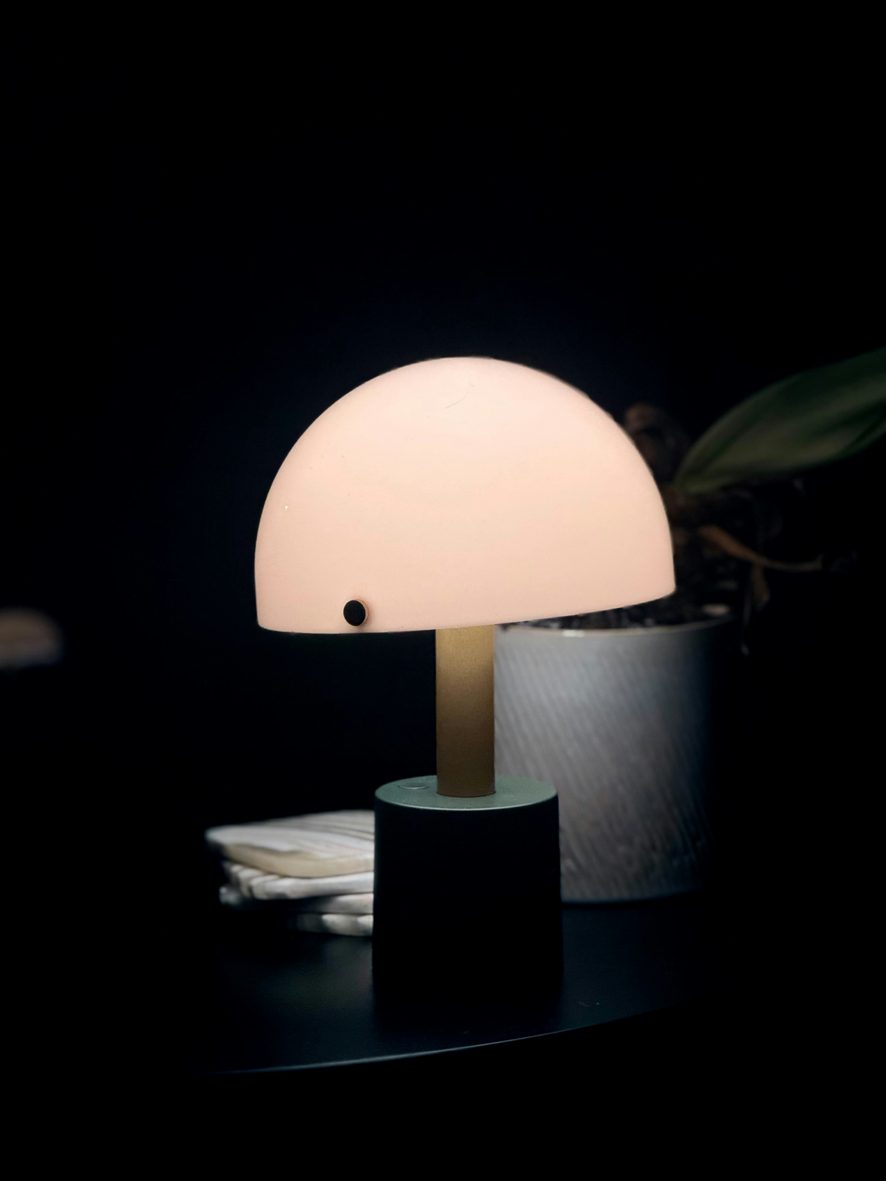 A softly glowing modern table lamp in a dark interior, surrounded by deep shadows and subtle greenery. Warm ambient light creates a quiet, moody atmosphere.