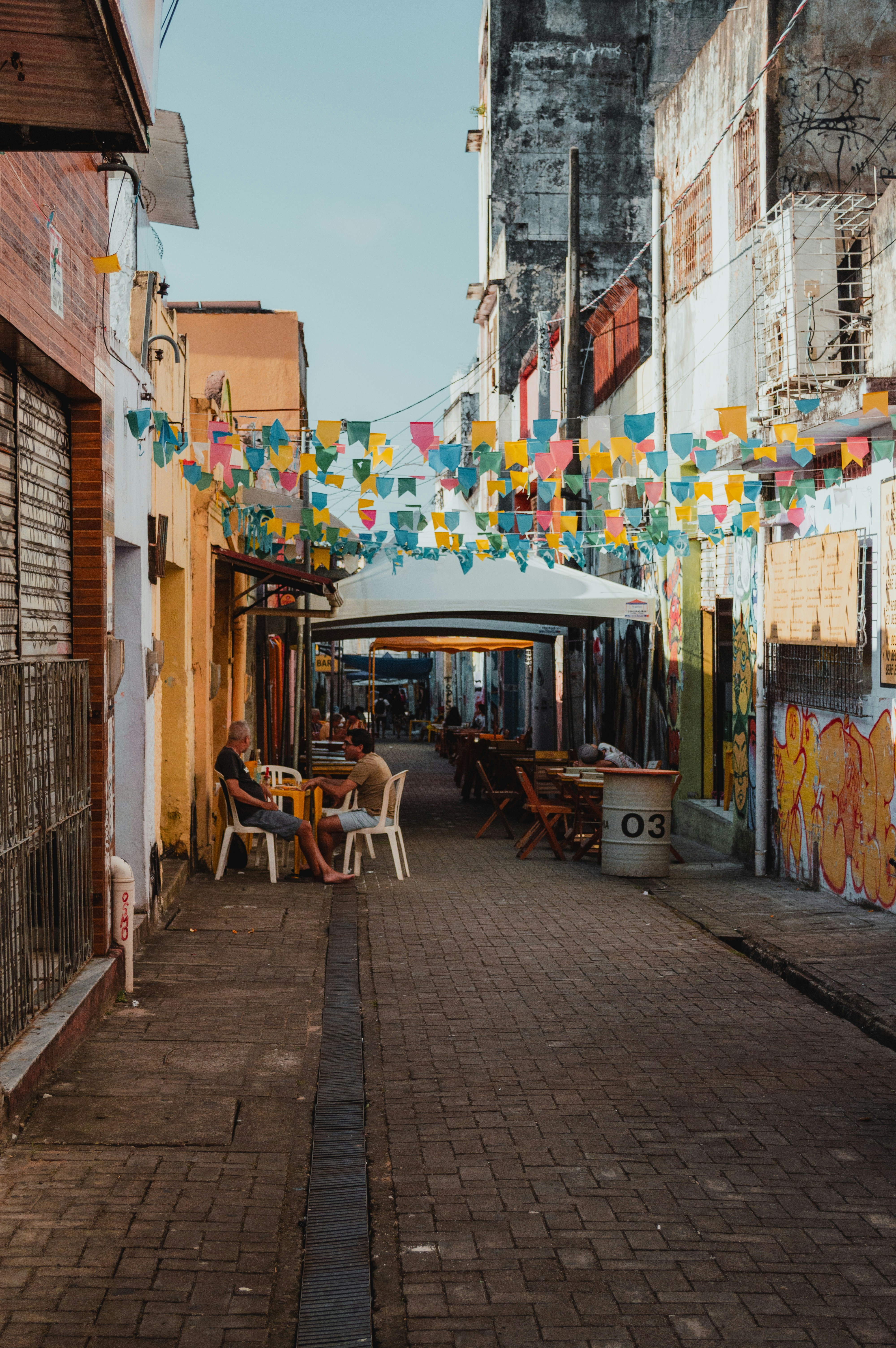 Colorful Street Corner: Dappled Light, People, and Picnic Tables Under Festive Banners