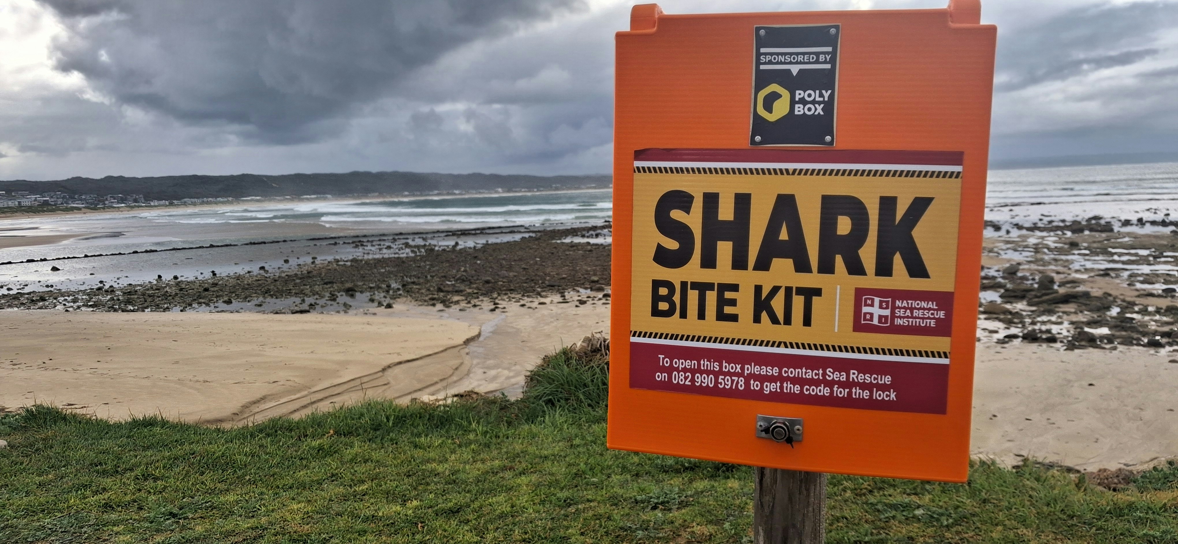 An orange sign reading "SHARK BITE KIT" on a post overlooking a sandy beach.