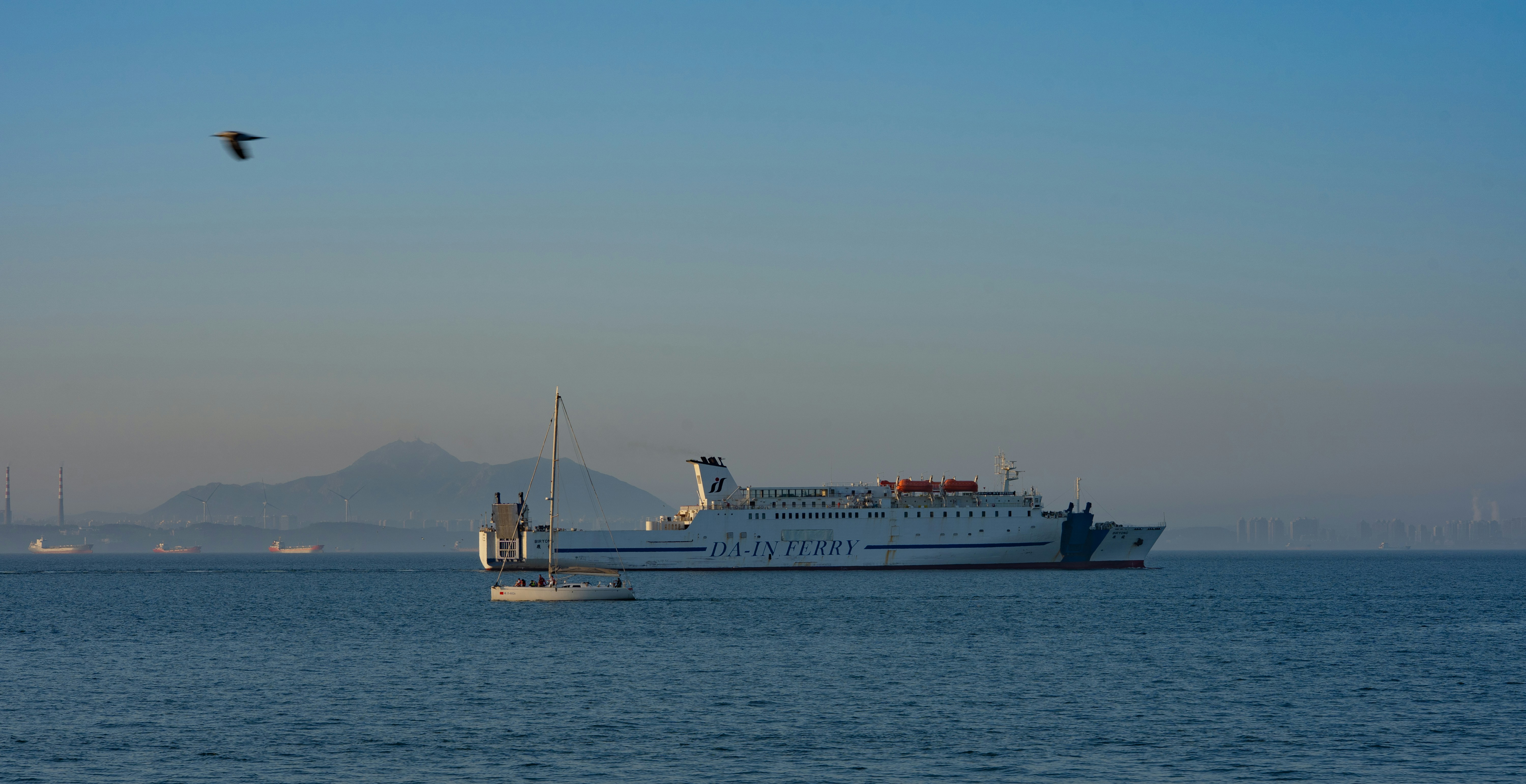 A calm seascape with a passenger ferry near the horizon and a lone sailboat in the foreground under a clear sky.