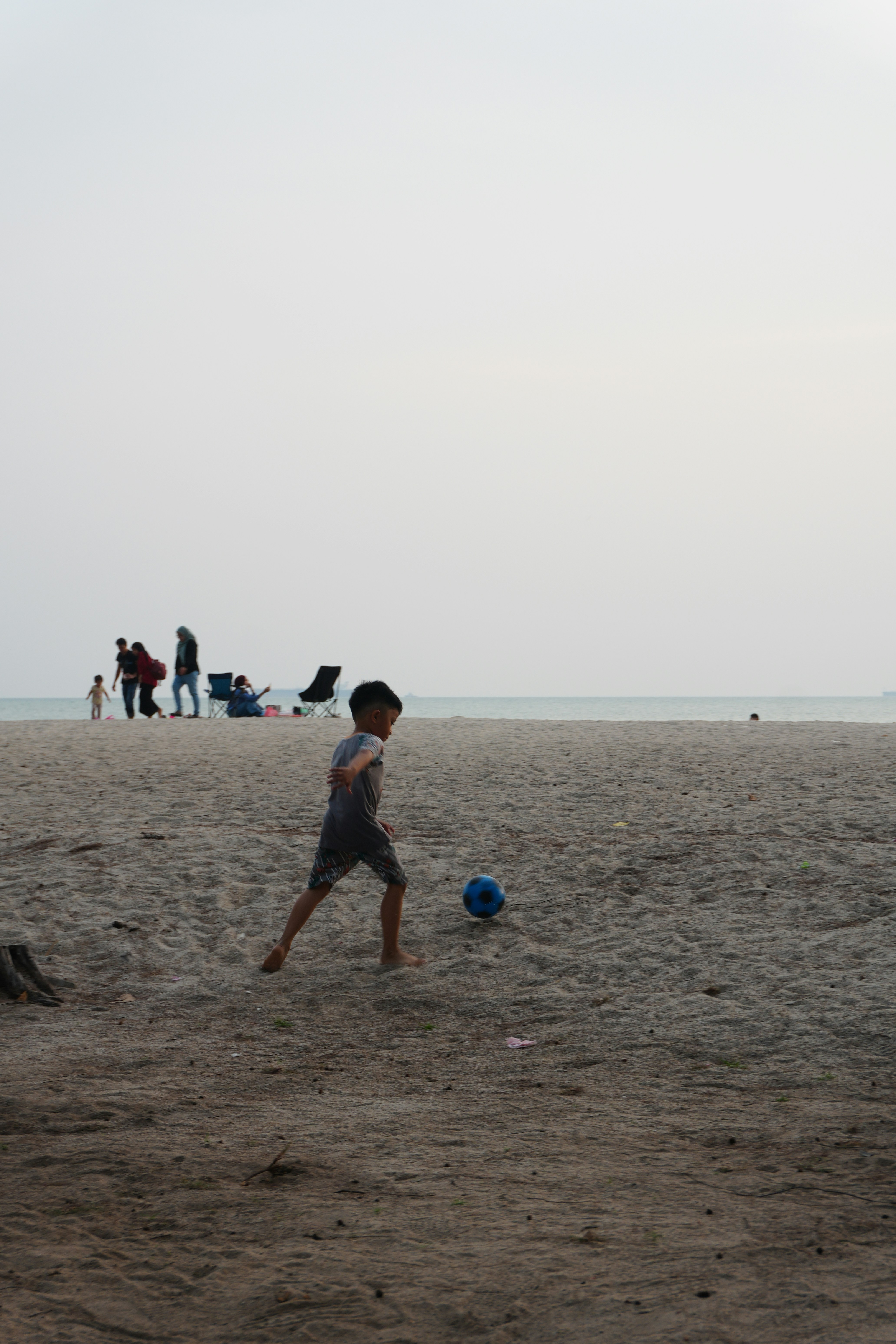 Sunlit beach scene featuring a child running after a blue ball, with a group of people and calm sea in the background.