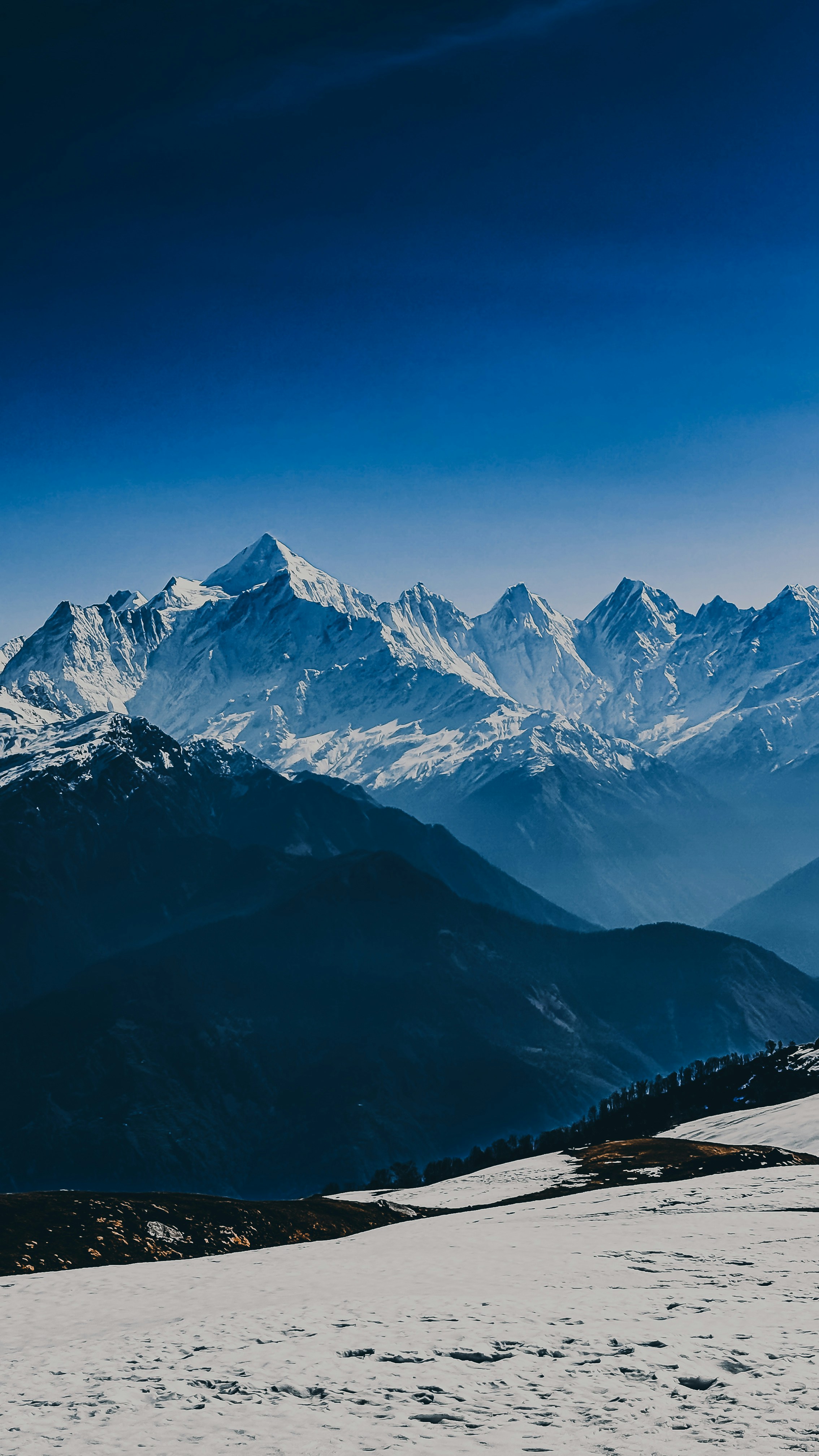 Snow-dusted alpine peaks rise under a deep blue sky, with foreground ridges fading into shadowed valleys. A tranquil landscape emphasizing vast scale and crisp contrast.