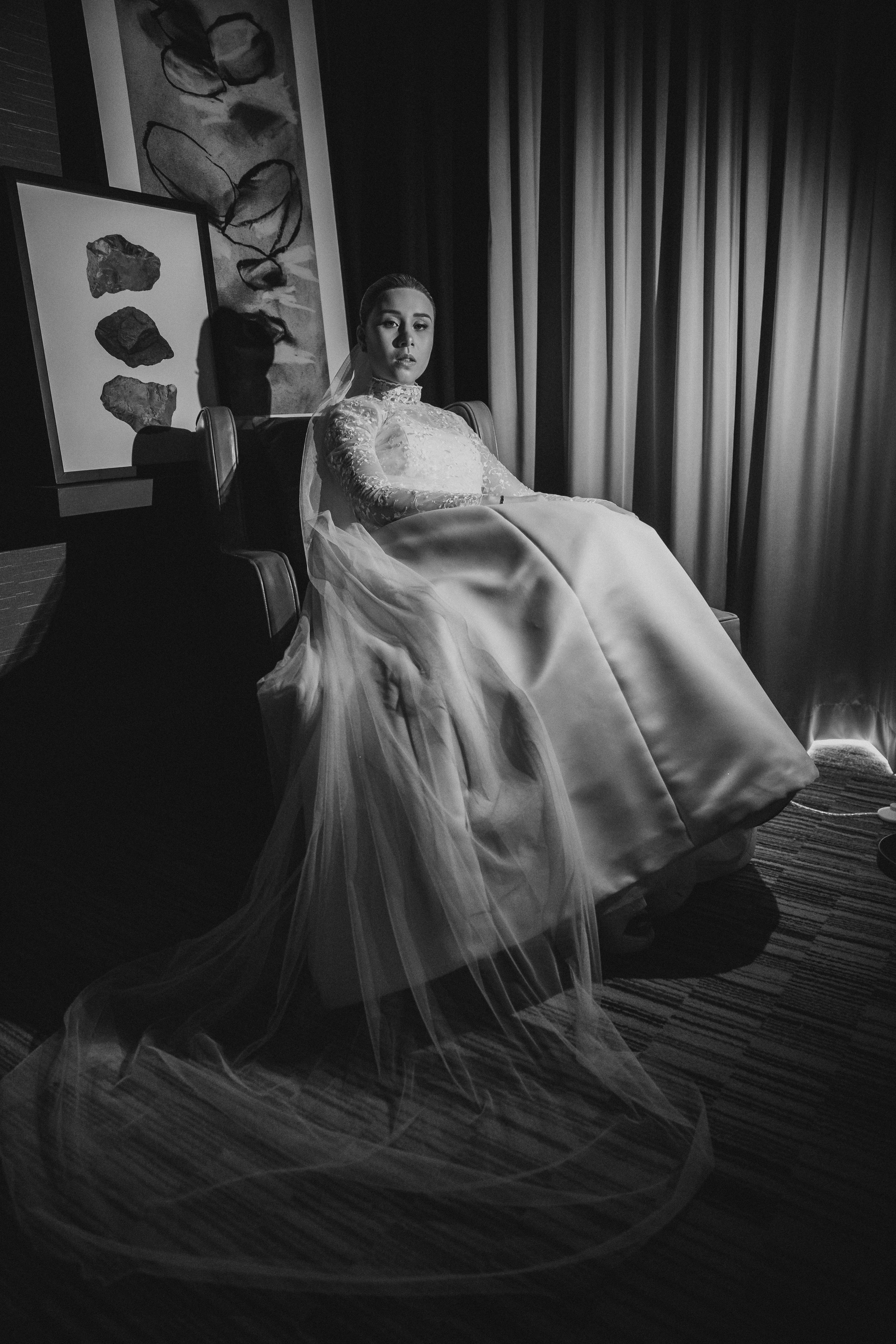 A woman in a flowing wedding gown sits on a velvet couch in a dimly lit gallery, the fabric cascading around her as framed artworks line the wall behind. A monochrome, painterly scene focusing on quiet drama.