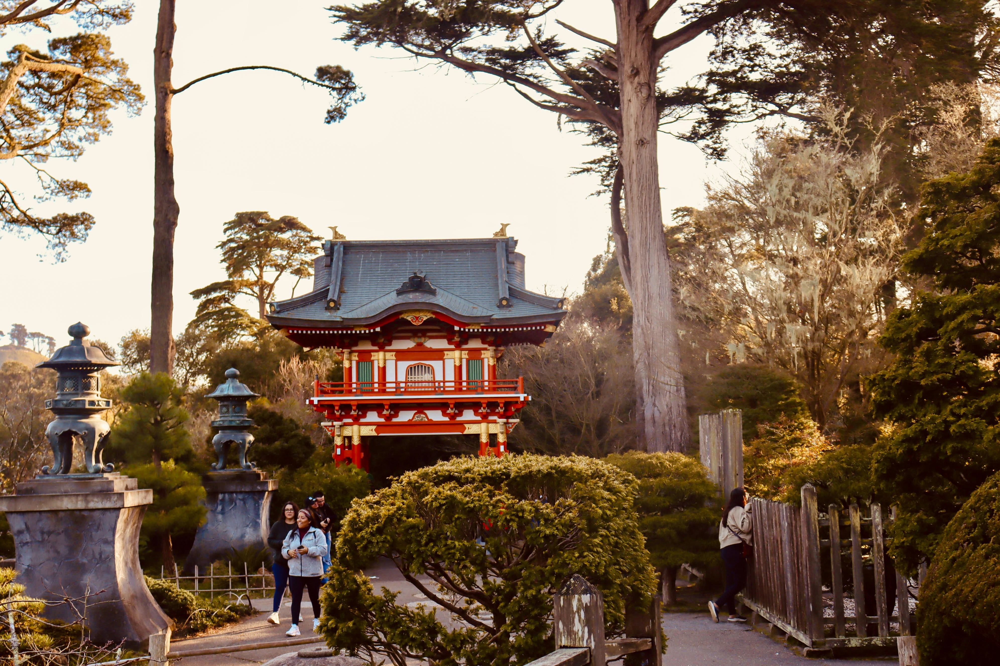 A tranquil Japanese garden with a red pavilion framed by tall trees and stone lanterns; visitors stroll along a winding path, suggesting a moment of quiet exploration.