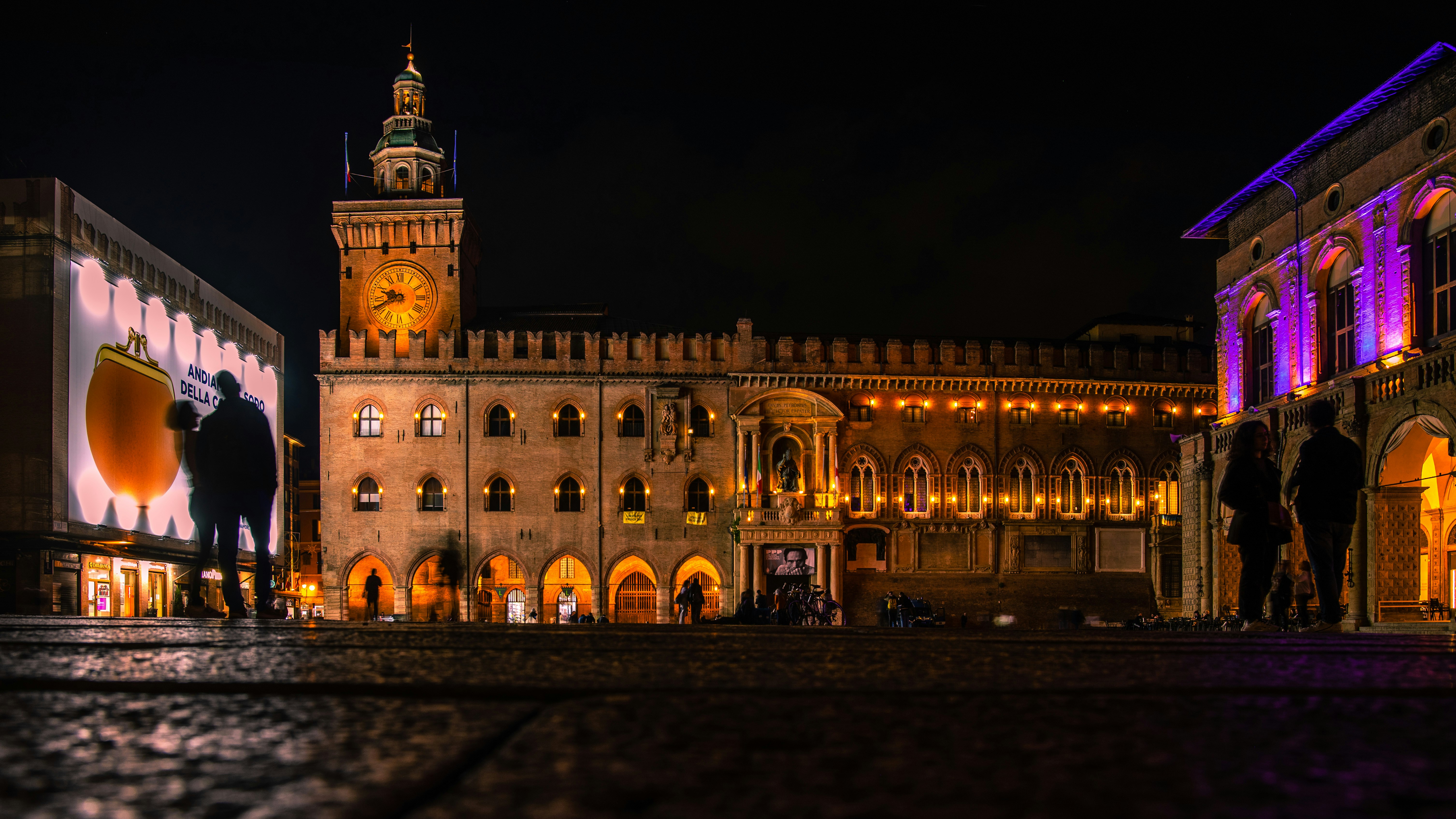 Piazza Maggiore in Bologna, Italy, comes alive at night with illuminated historic buildings, colorful architectural lighting, and pedestrians in the square. A dramatic travel photograph showcasing Italian architecture and urban nightlife.