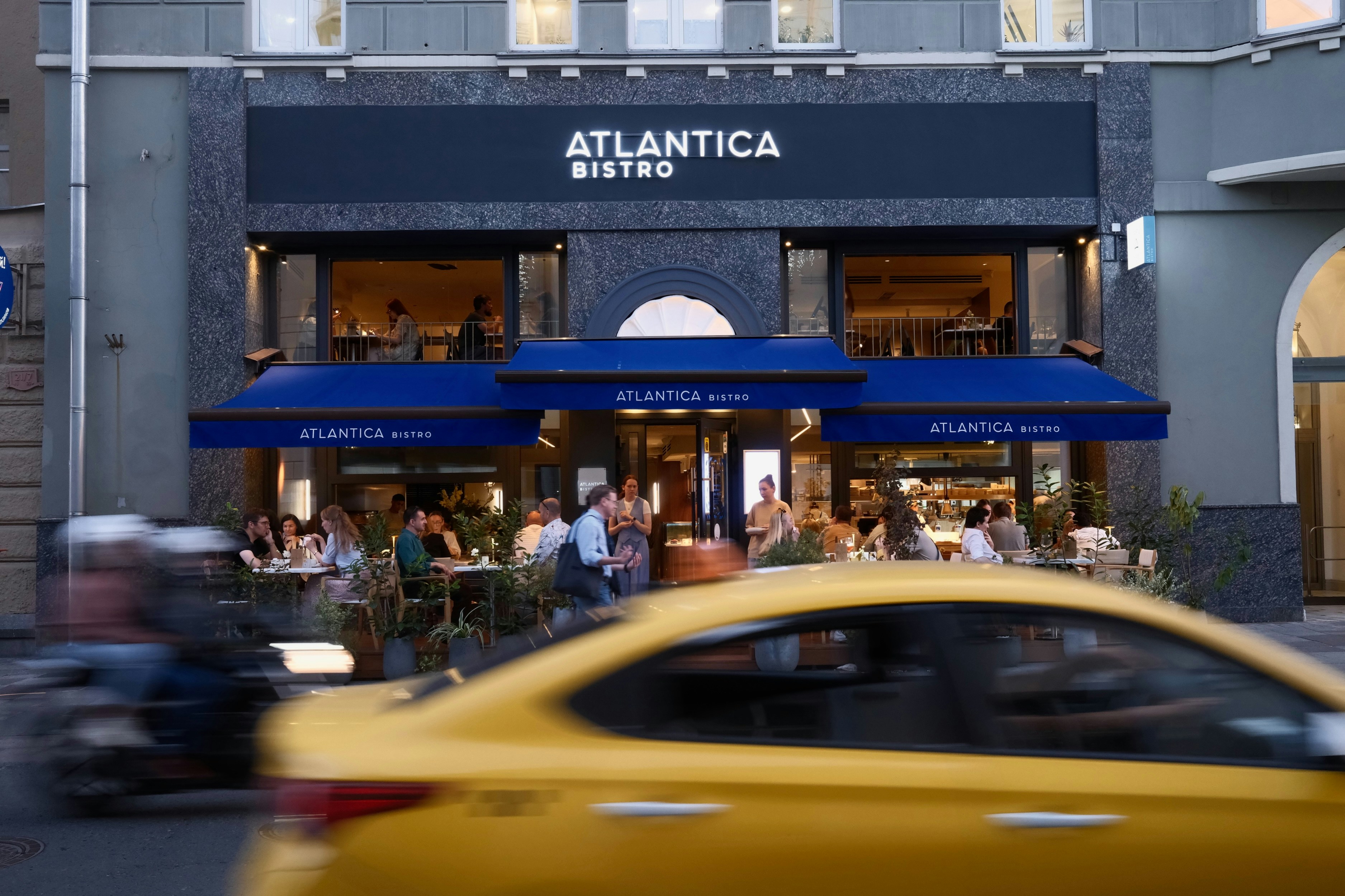 Energetic street view of Atlantica Bistro with diners under blue awnings, blurred yellow taxi in motion in foreground.