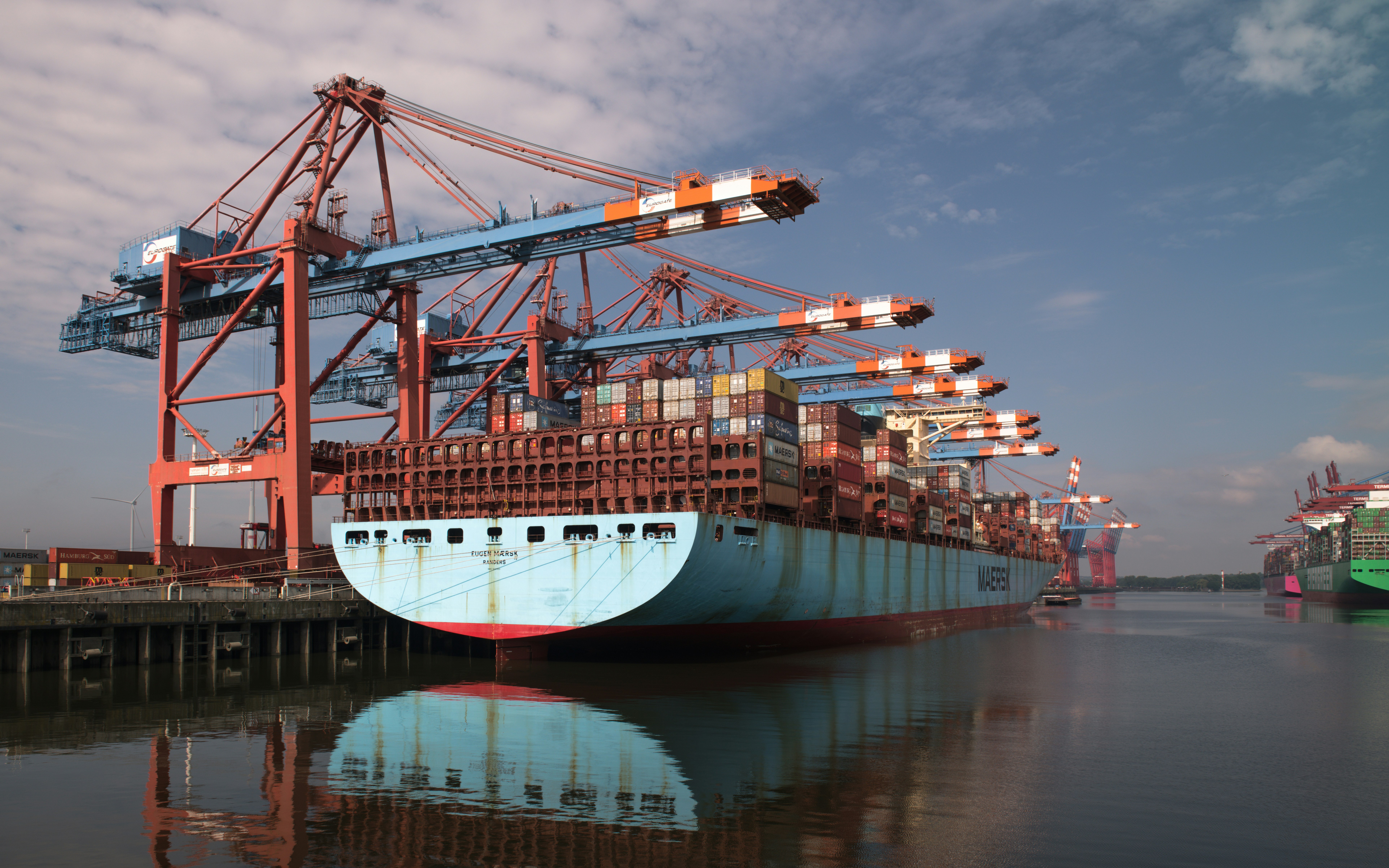 A large container ship docked at a port with red cranes overhead.