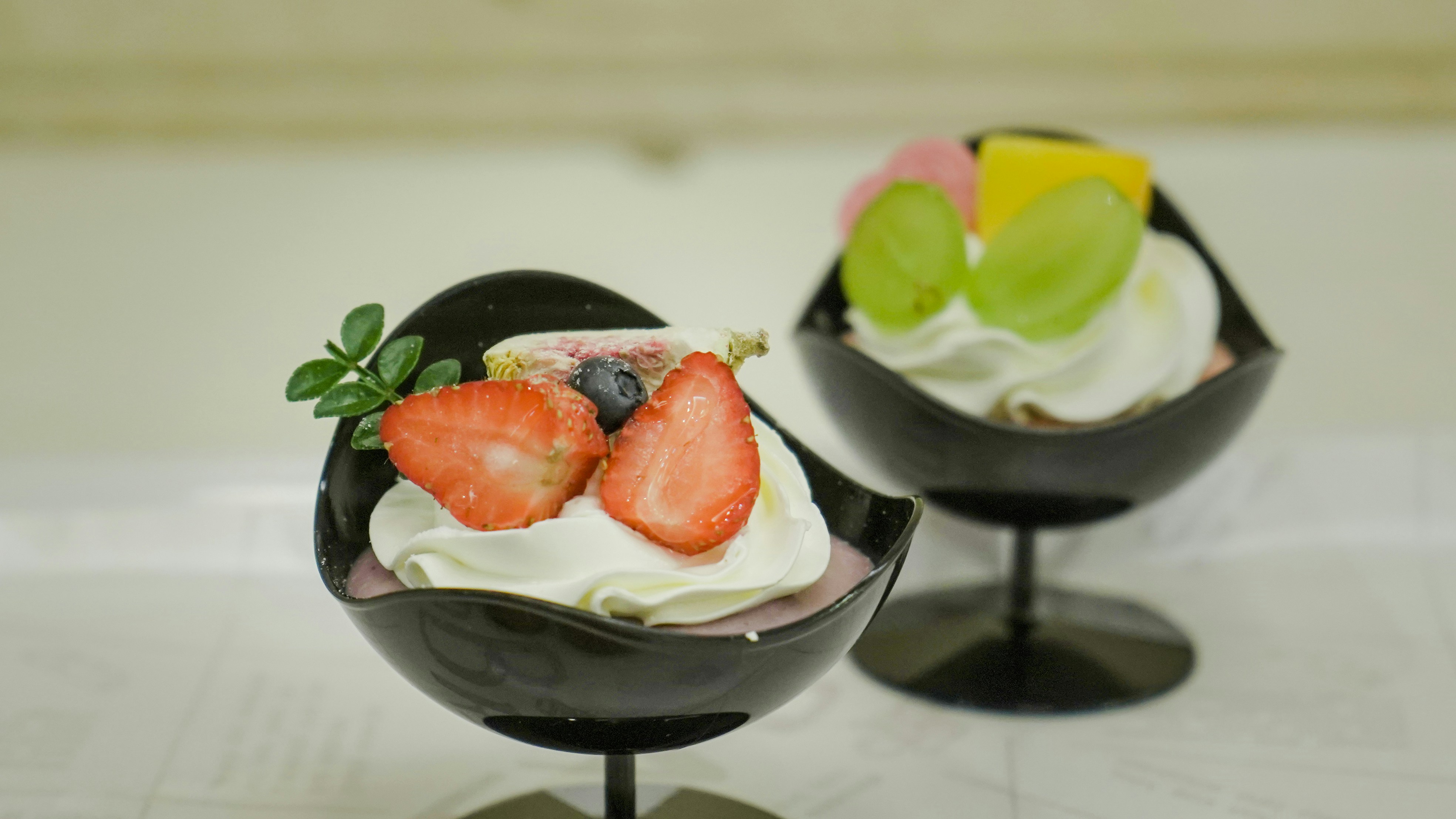 Two glossy dessert bowls on a light surface, topped with whipped cream and fresh fruit slices. A close, minimal setting highlighting presentation and color contrast.